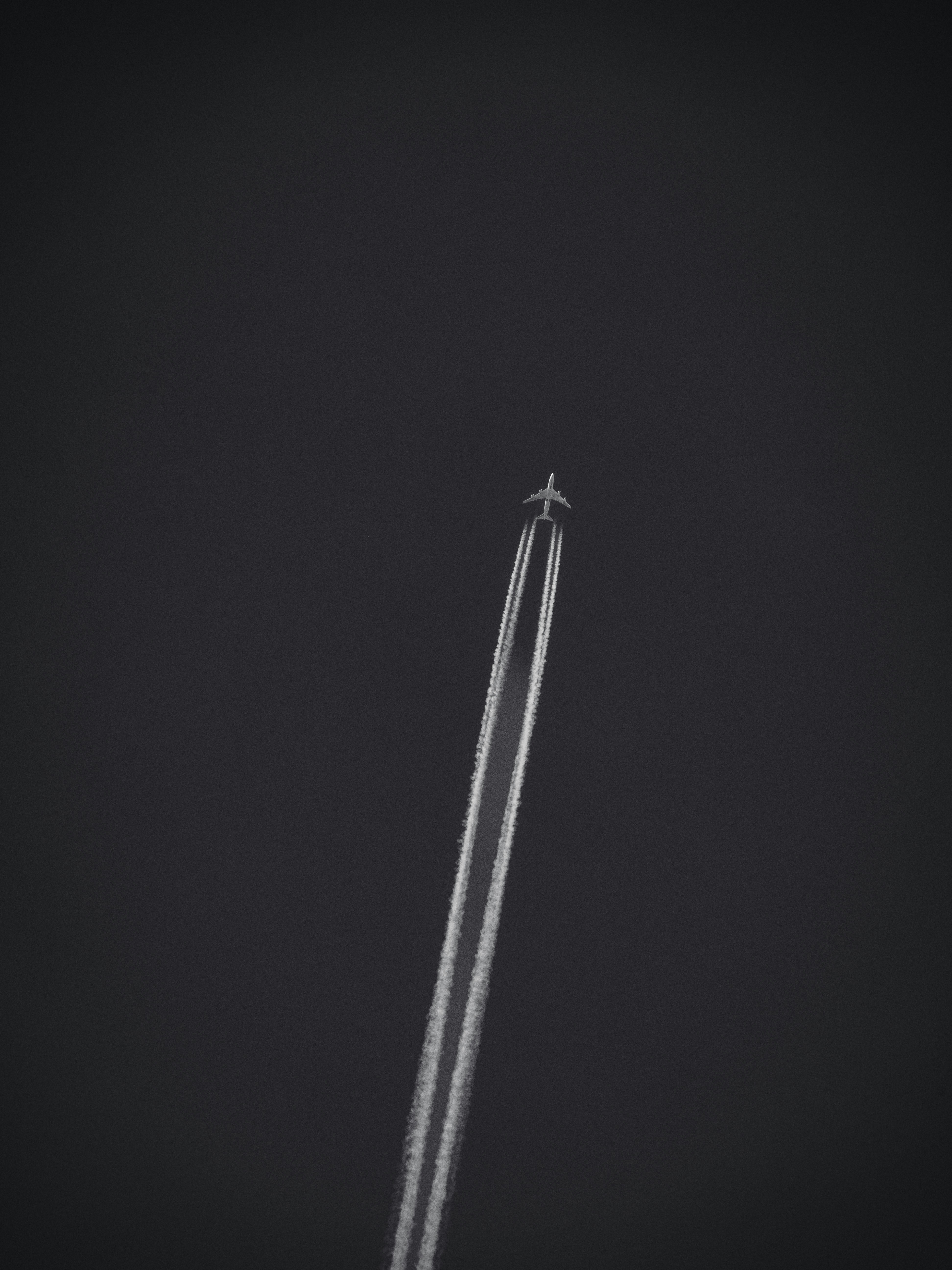 A minimal monochrome photograph of an airplane high in the sky, leaving two white trails behind. A calm and cinematic image about movement, distance, and silence. | Airplane leaving contrails against a dark sky