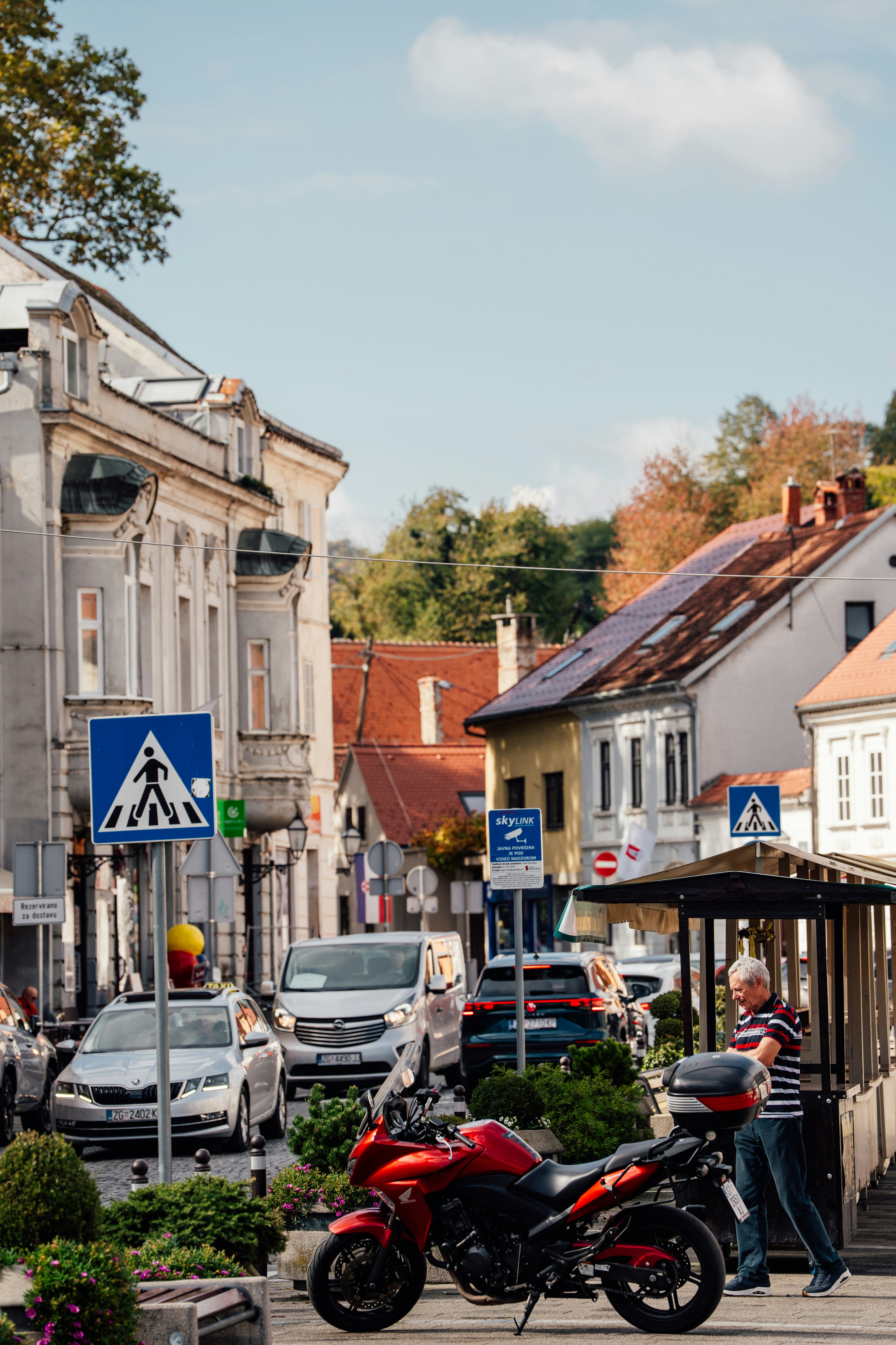 A vibrant street scene featuring a red motorcycle parked in the foreground, with pedestrians and vehicles amidst charming architecture and traffic signs.