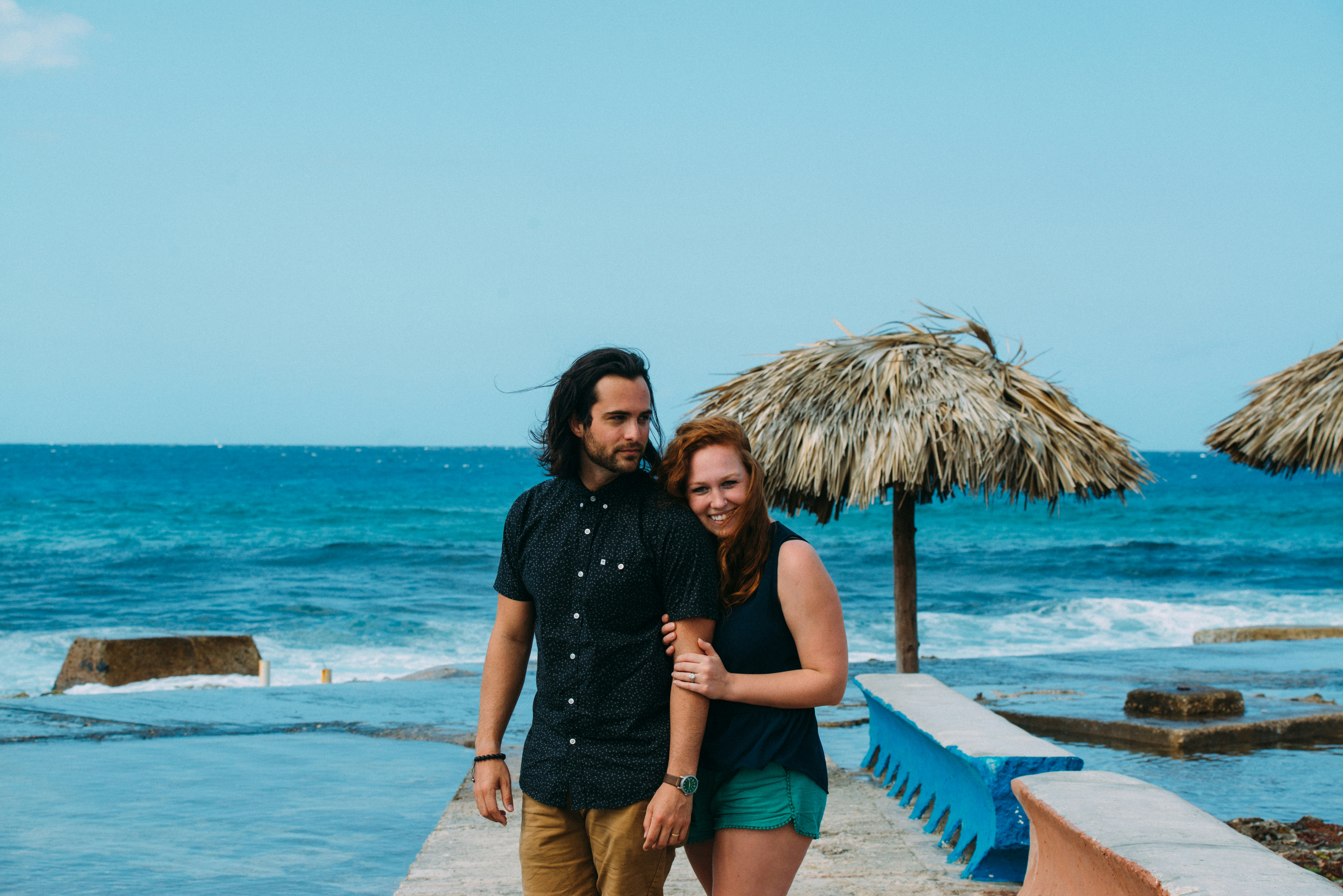 Couple enjoying a joyful moment by the ocean, framed by palm thatched umbrellas and vibrant blue waters.