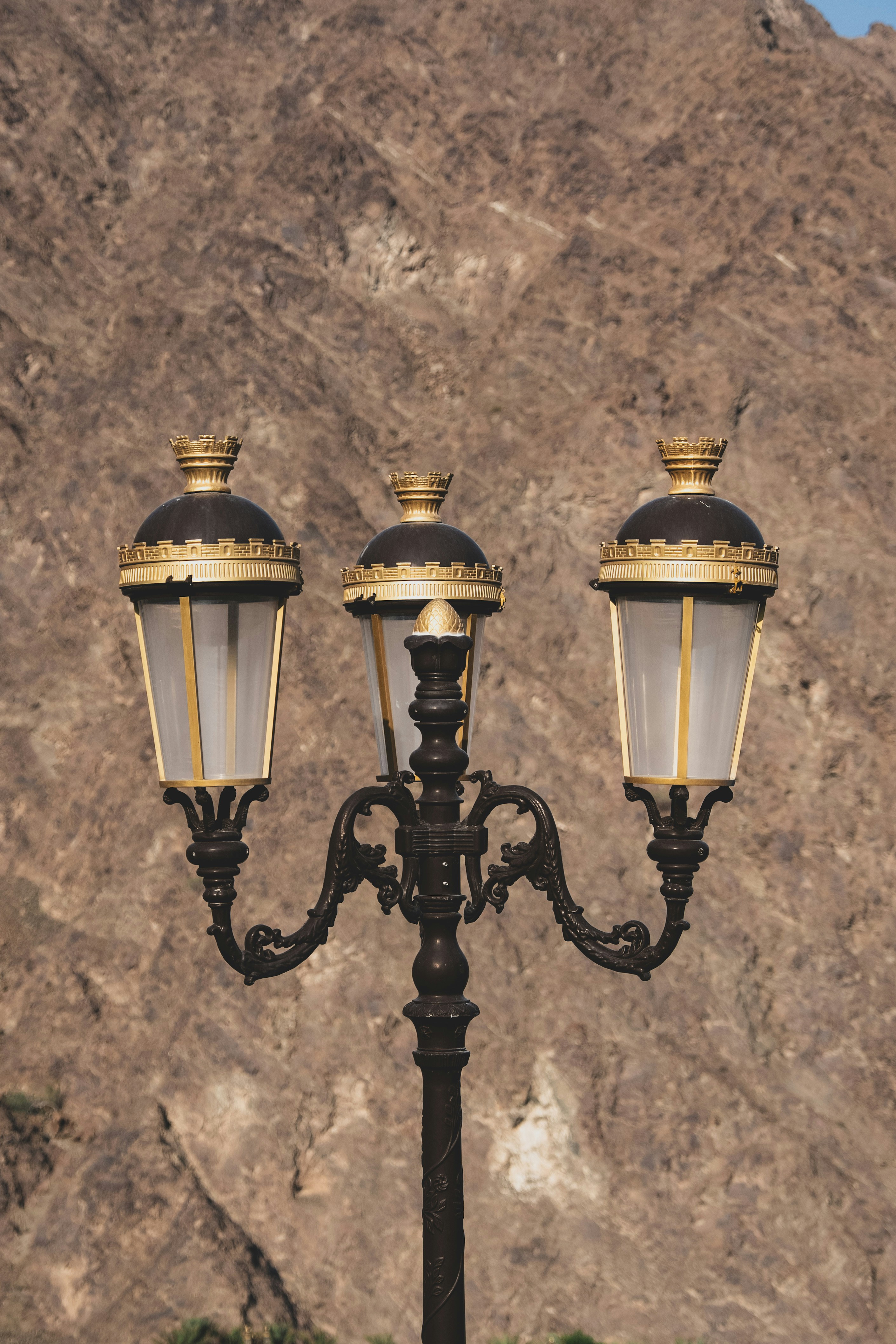 Ornate street lamp with three glass shades set against a rugged mountain backdrop. The design combines classic elegance with natural beauty.