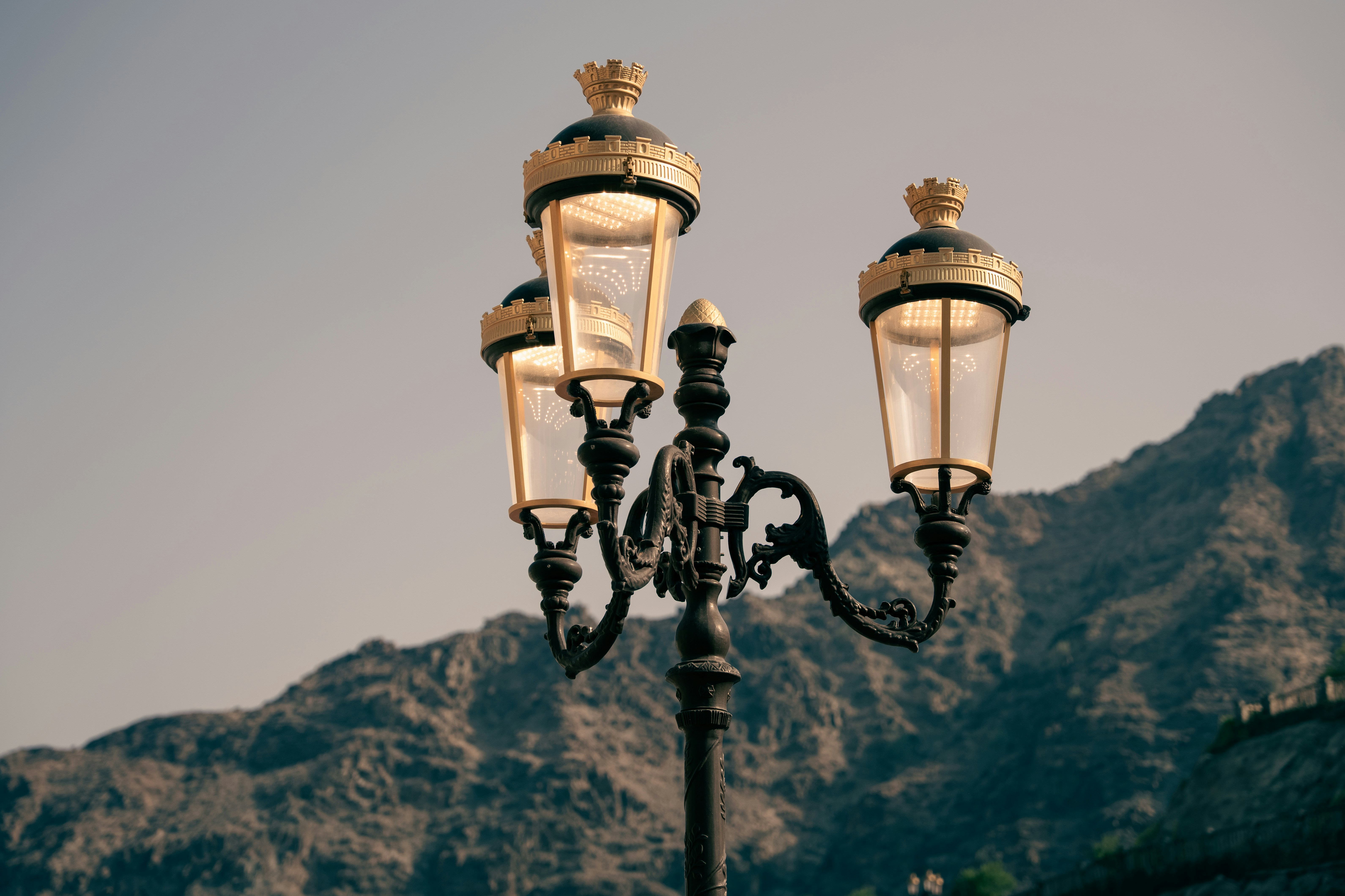 When even streetlights become works of art! Details that transform landscapes into visual poetry. | Ornate street lamp with three lights against mountains.