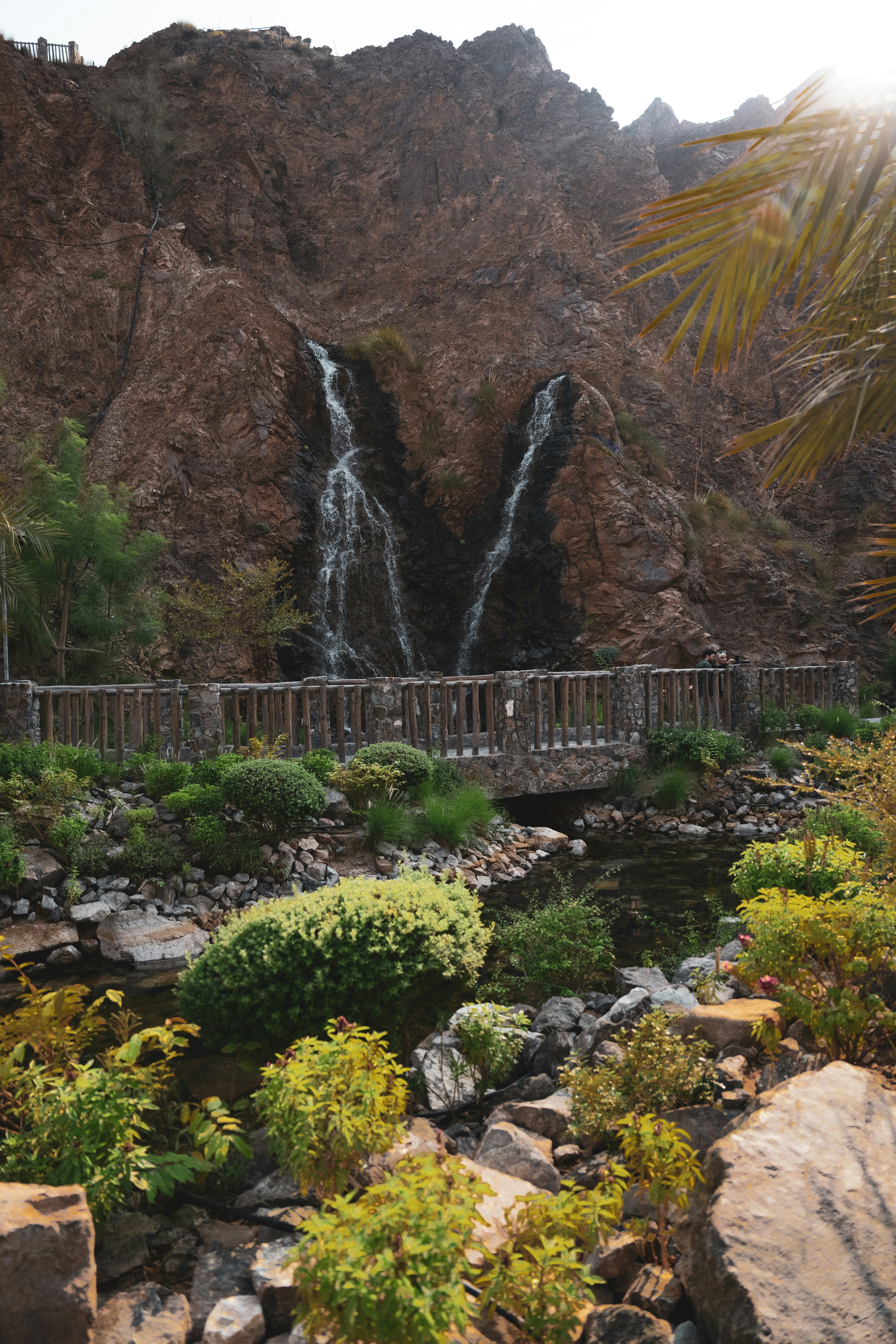 Waterfall cascading down rocky cliffs surrounded by lush greenery and a wooden bridge. A tranquil scene of nature's harmony.
