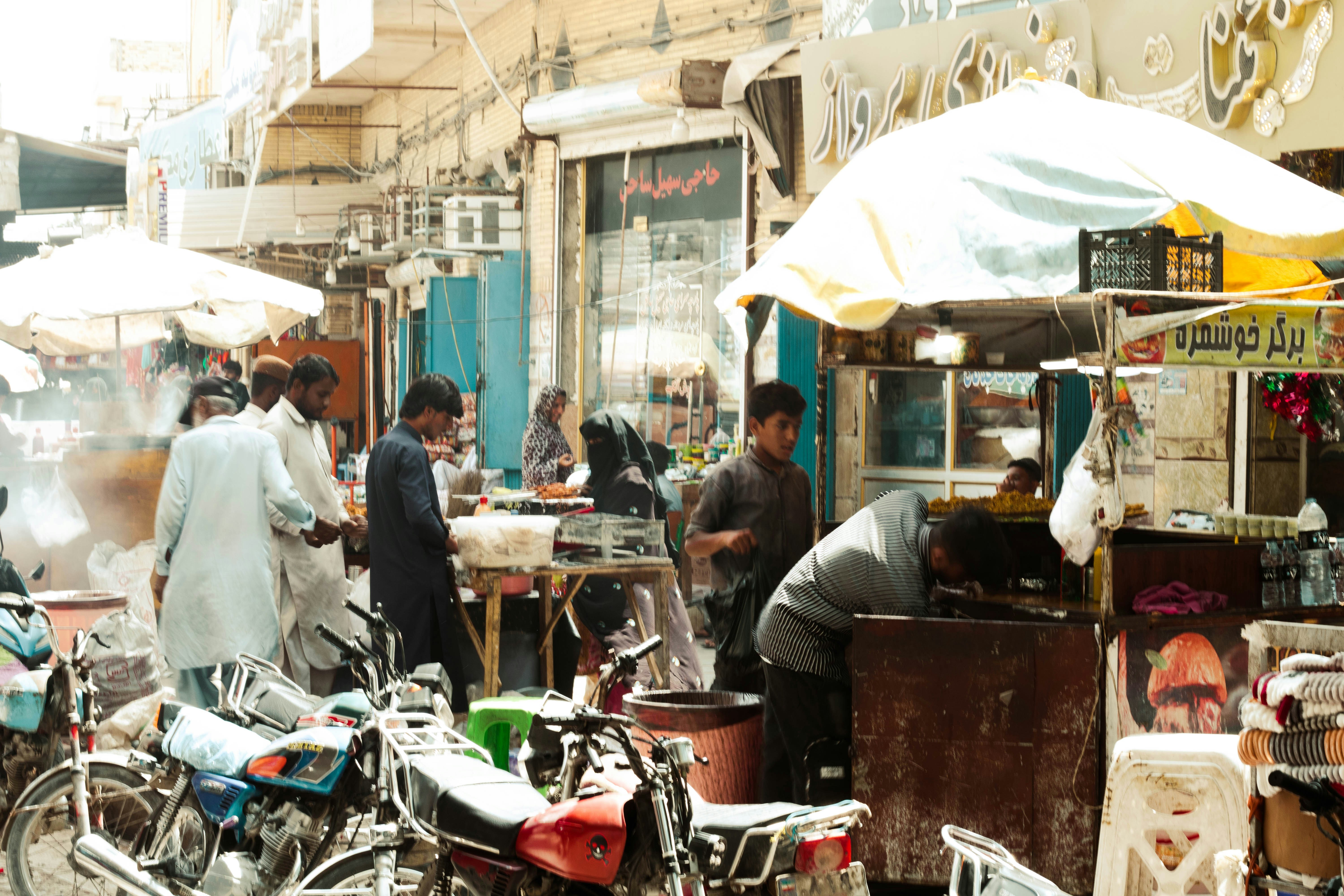 Busy street market scene with vendors and customers interacting amidst colorful bicycles and food stalls.