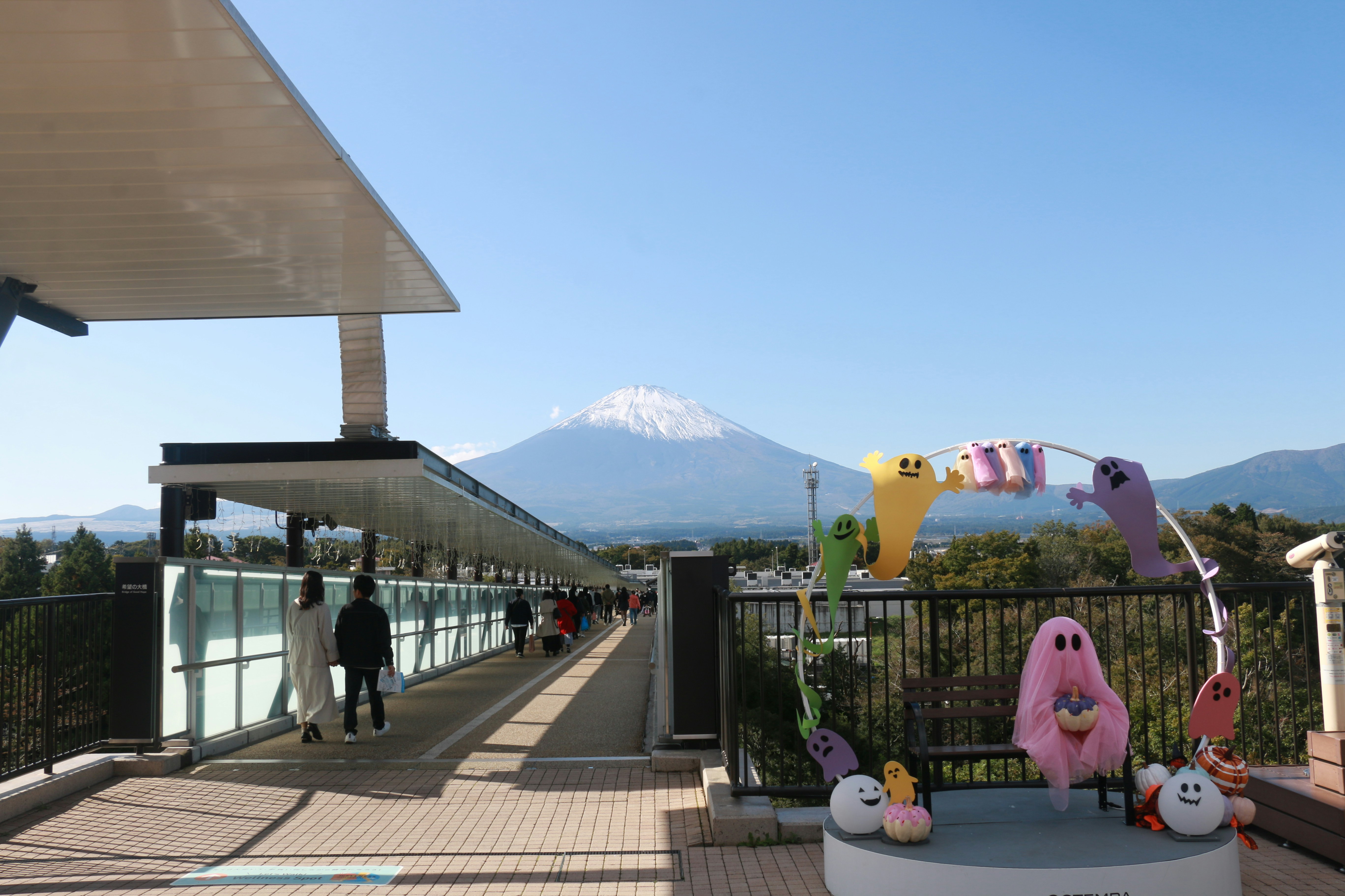 Colorful ghost figures adorn a bench overlooking a serene view of Mount Fuji. The scene captures a blend of playful art and natural beauty.