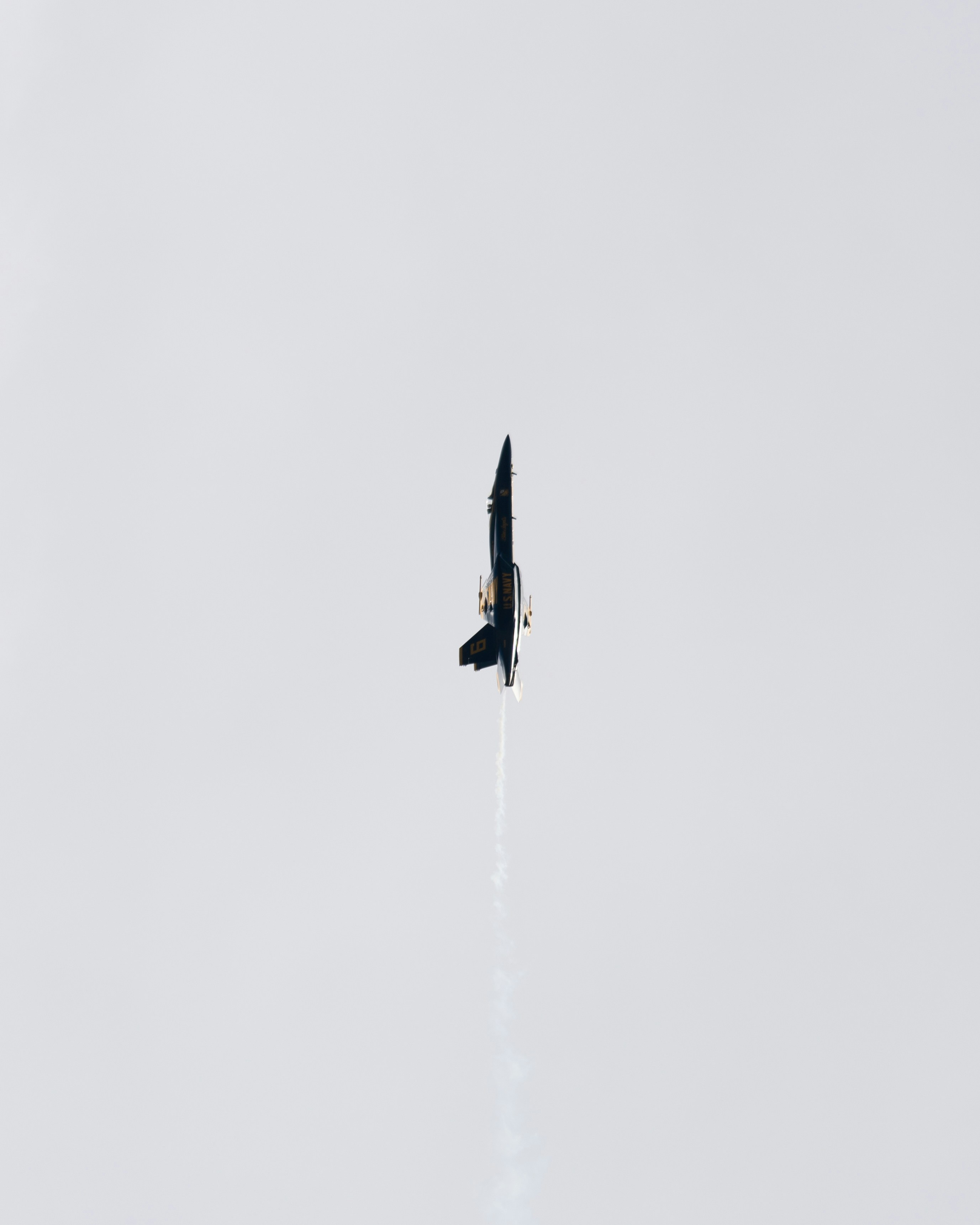 Fighter jet performing a vertical climb against a pale sky, leaving a trail of vapor behind. 