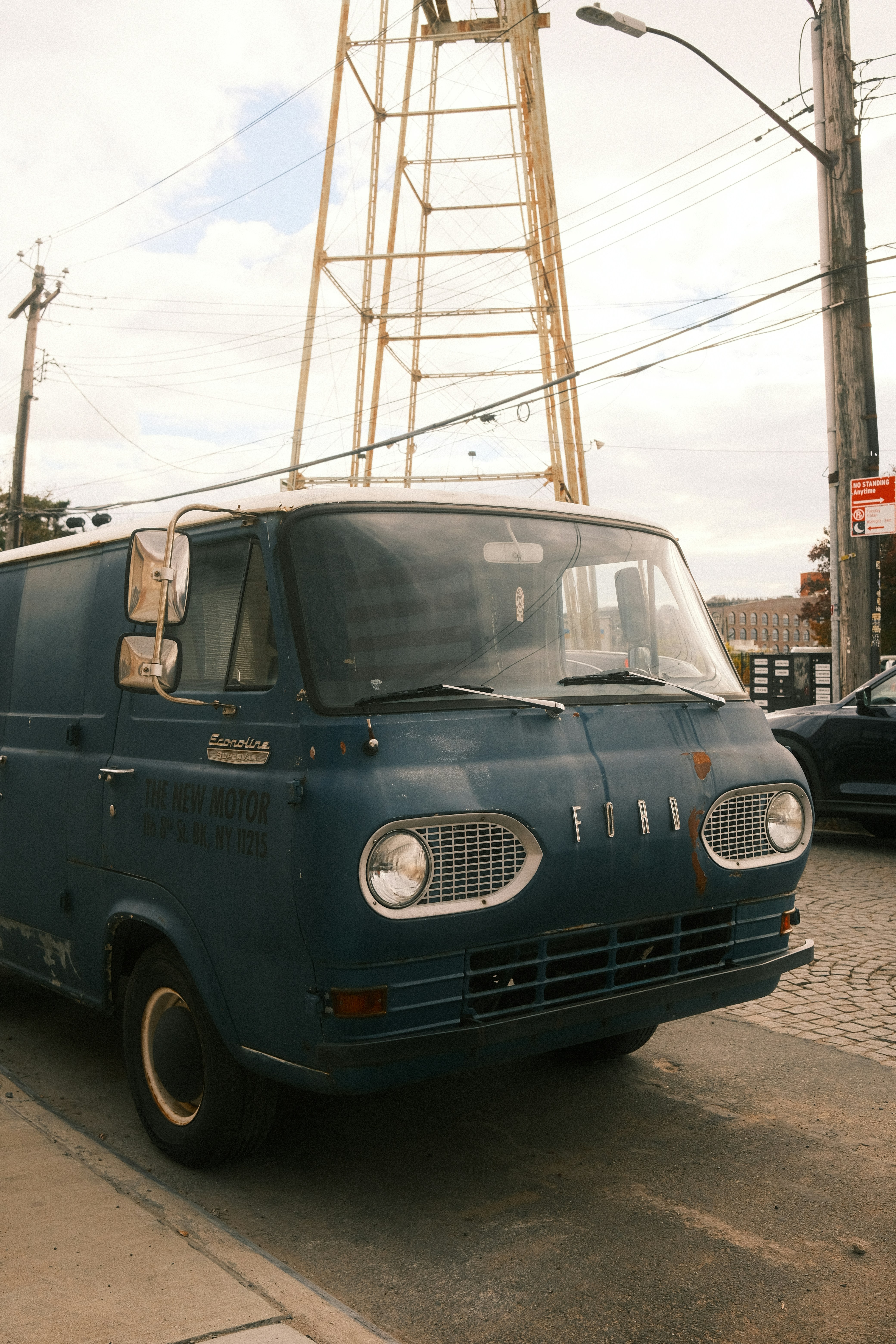 A vintage blue ford van parked by a tall structure.