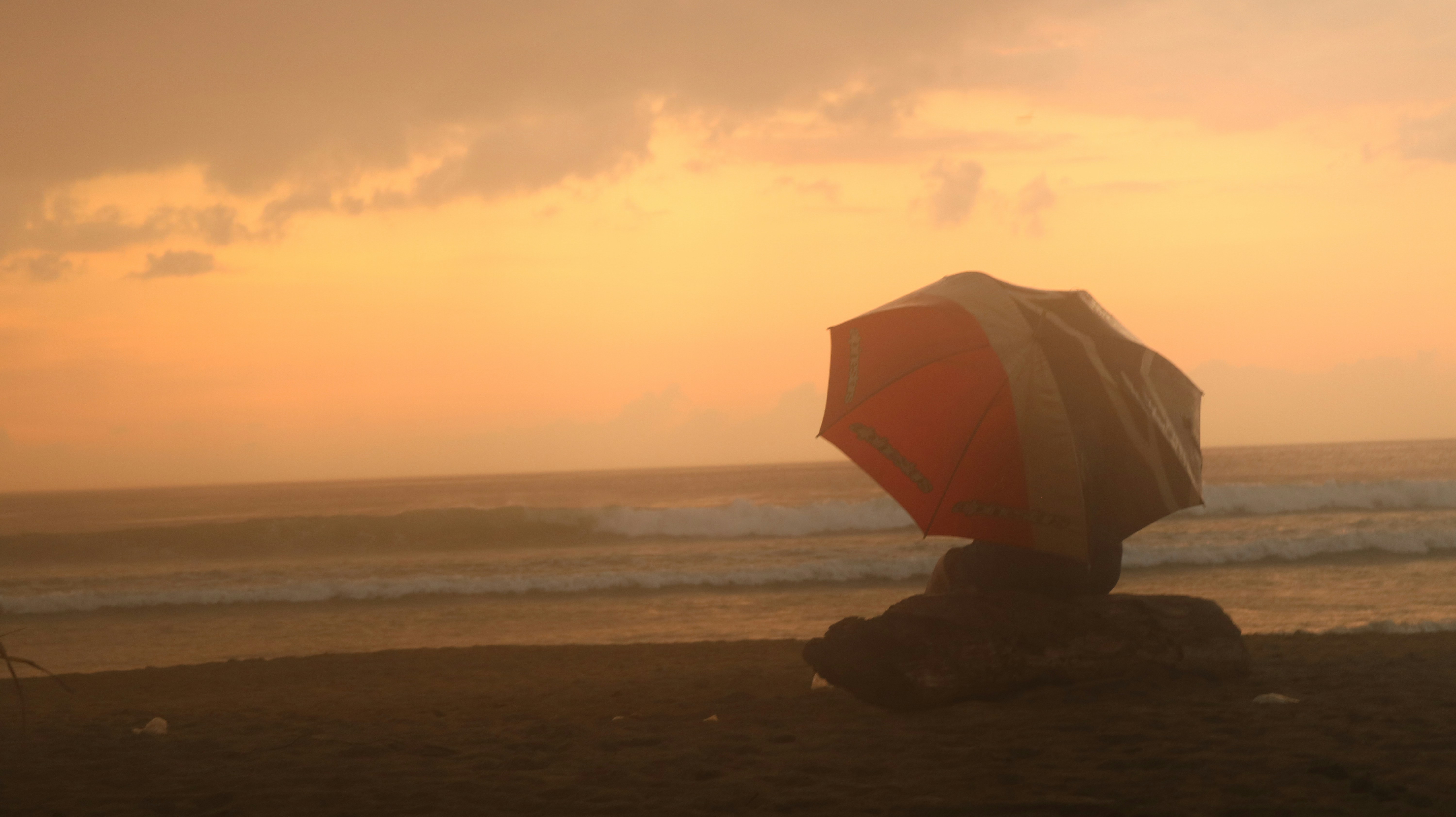 Vibrant orange umbrella perched on a rock, silhouetted against a golden sunset over the ocean waves.