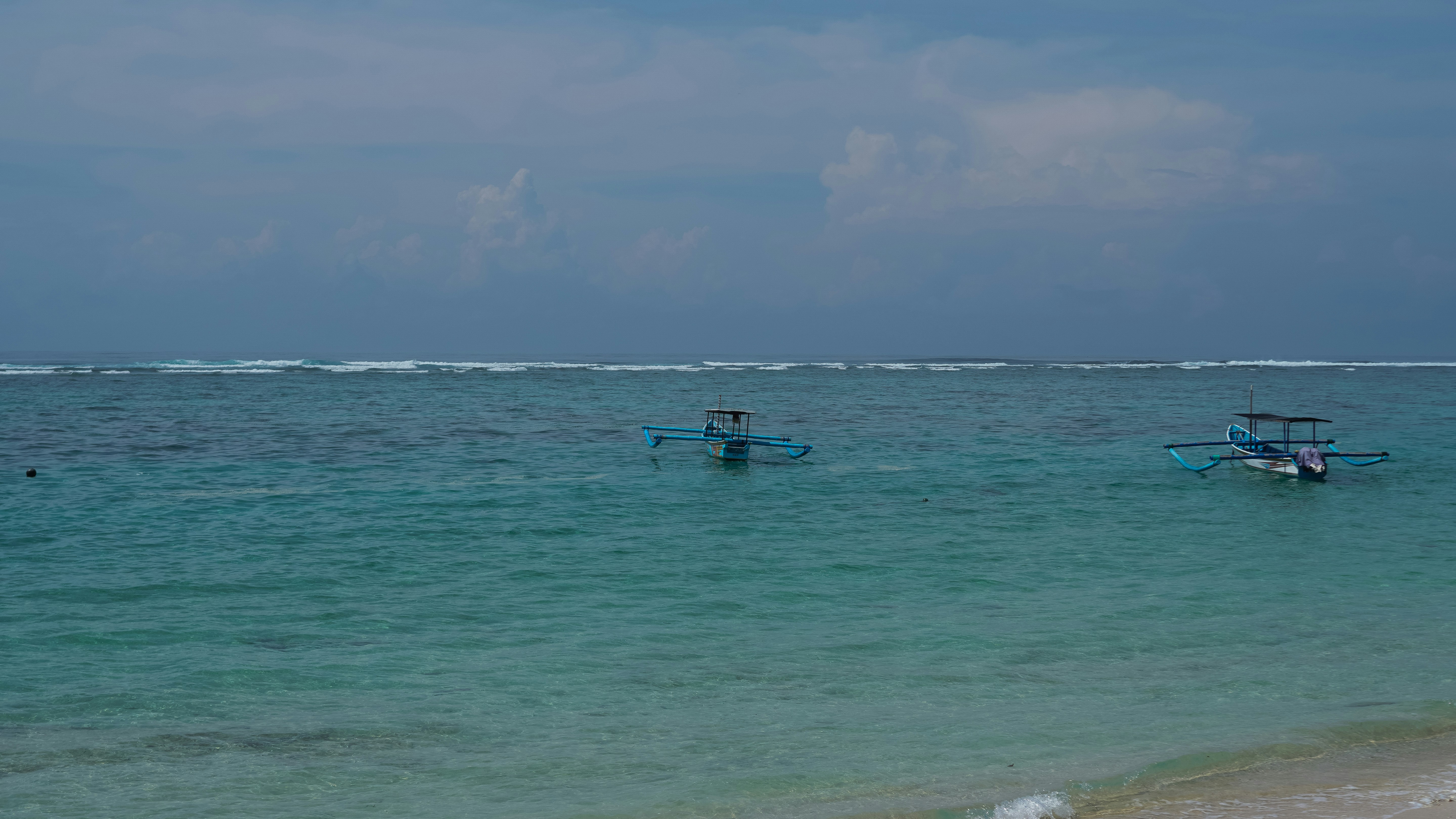 Two traditional boats anchored in calm turquoise waters under a cloudy sky. The scene captures the essence of coastal tranquility.