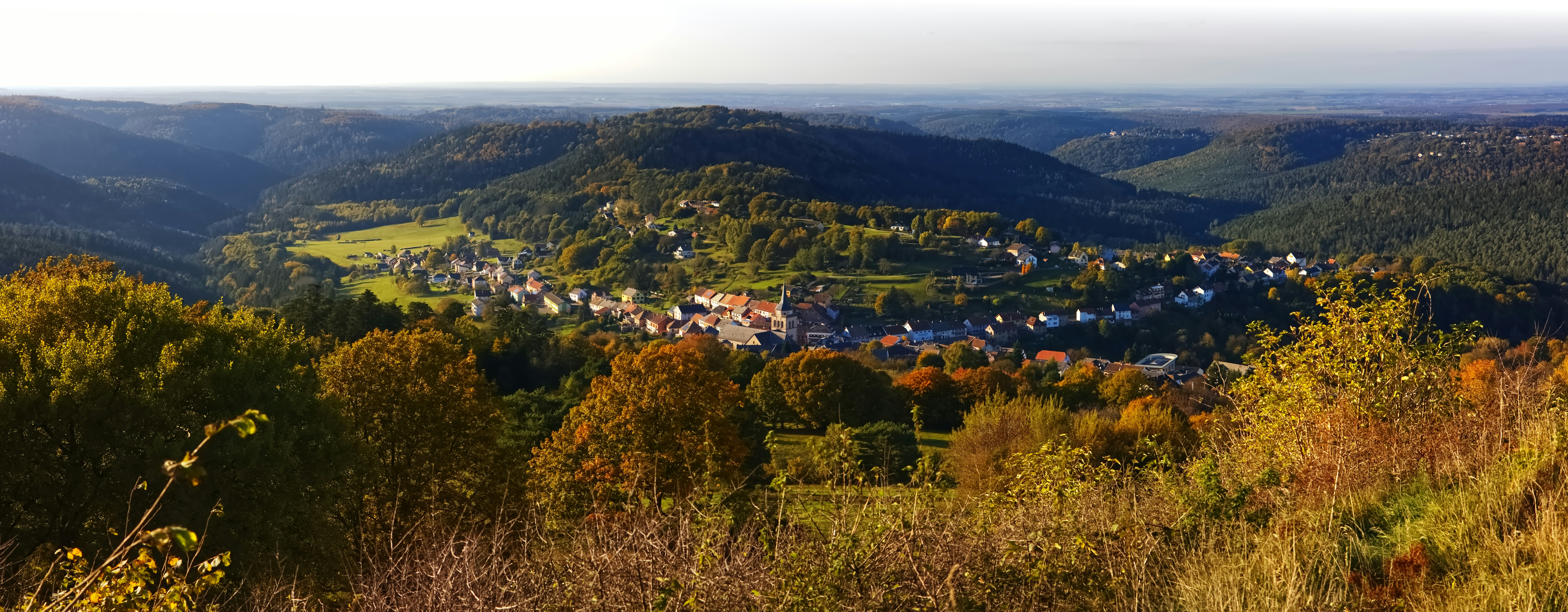 A panoramic view of a quaint village nestled among rolling hills and vibrant autumn foliage. The scene captures the serene coexistence of nature and human settlement.
