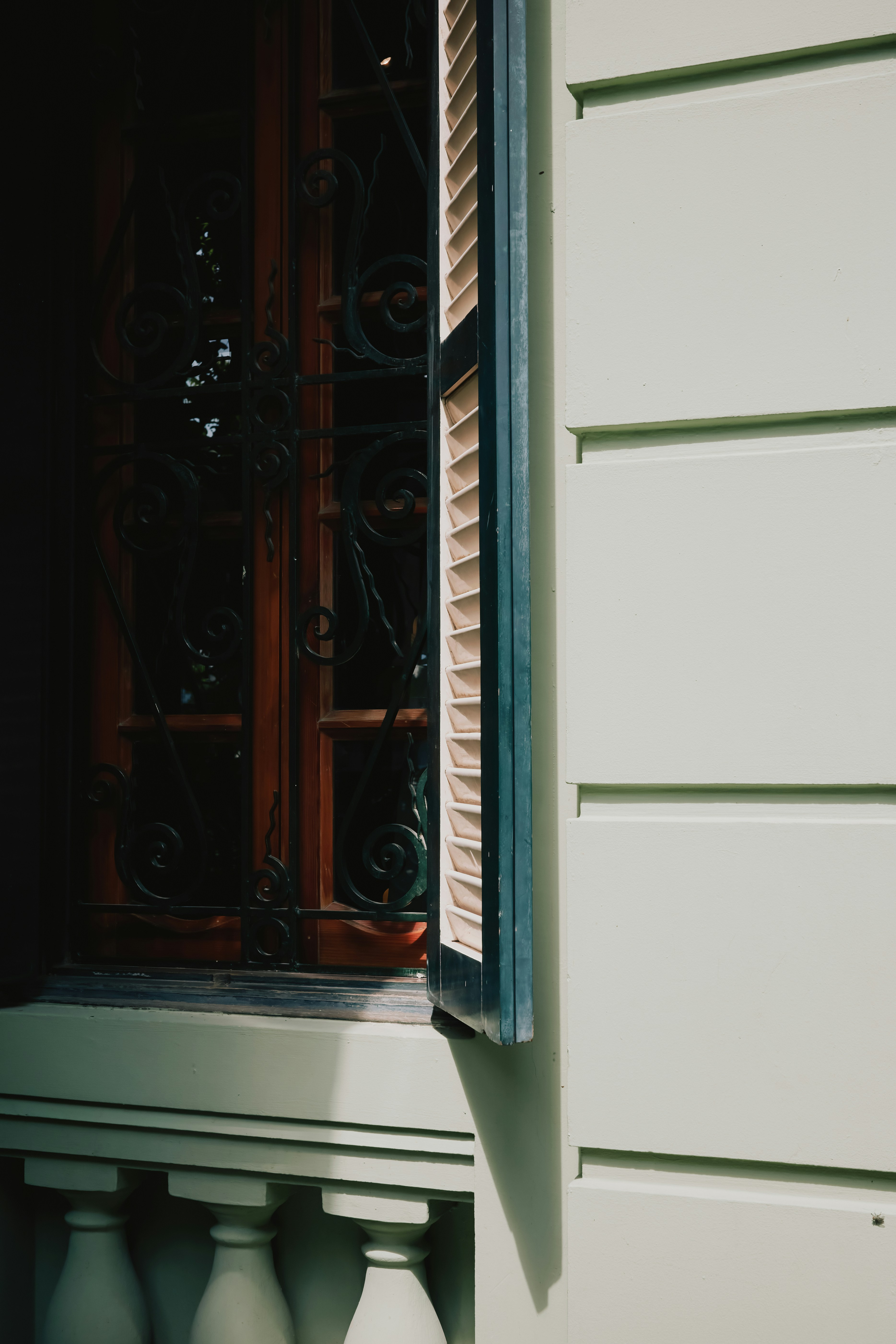 An open wooden window with louvered shutters and decorative black iron bars, set in a pale green exterior wall. Sunlight casts strong shadows on the building’s facade. | Open window with ornate ironwork and wooden door