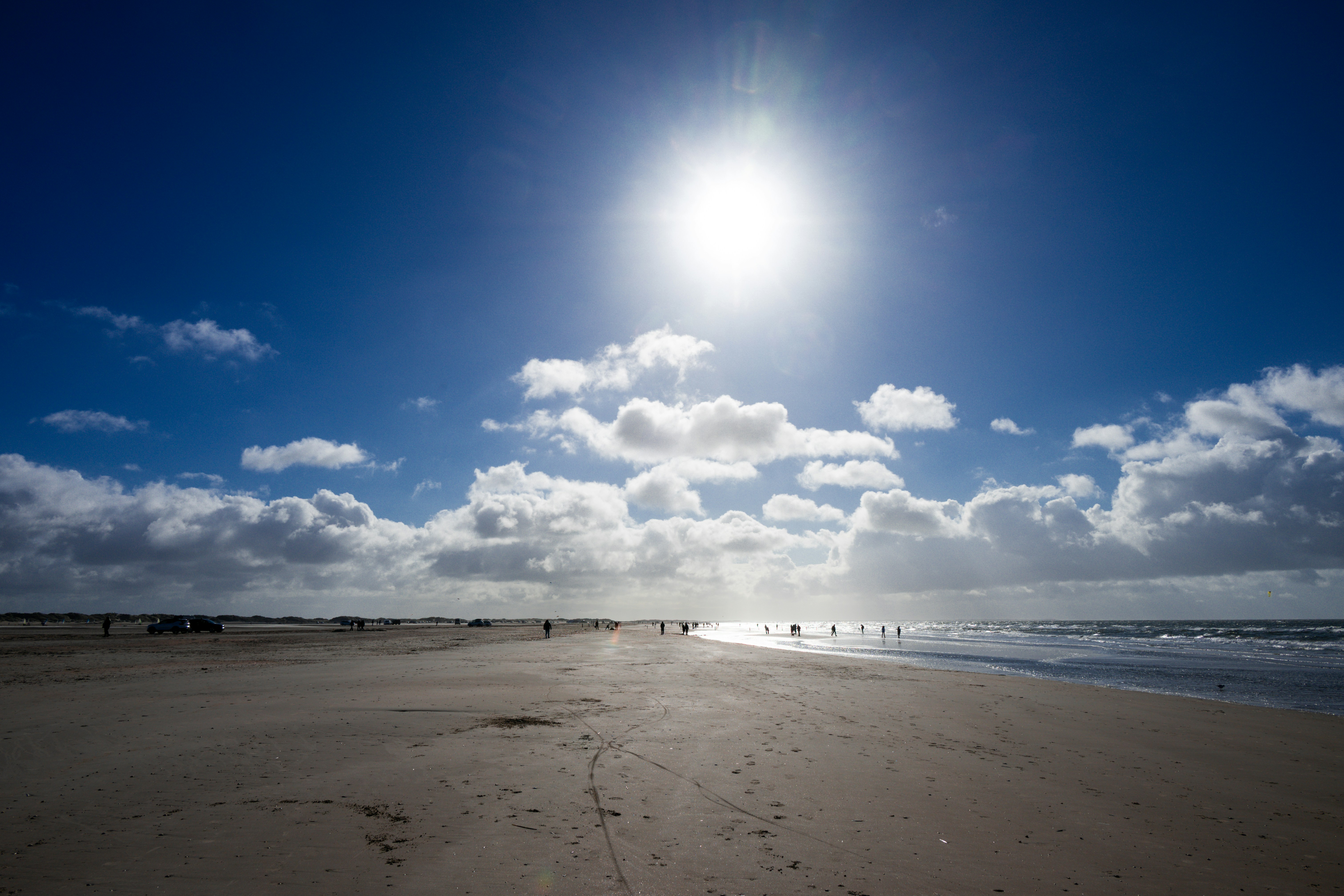 A serene beach scene under a bright sun, with scattered clouds reflecting the tranquil atmosphere. Footprints trace the sandy shore where distant figures enjoy the ocean breeze.