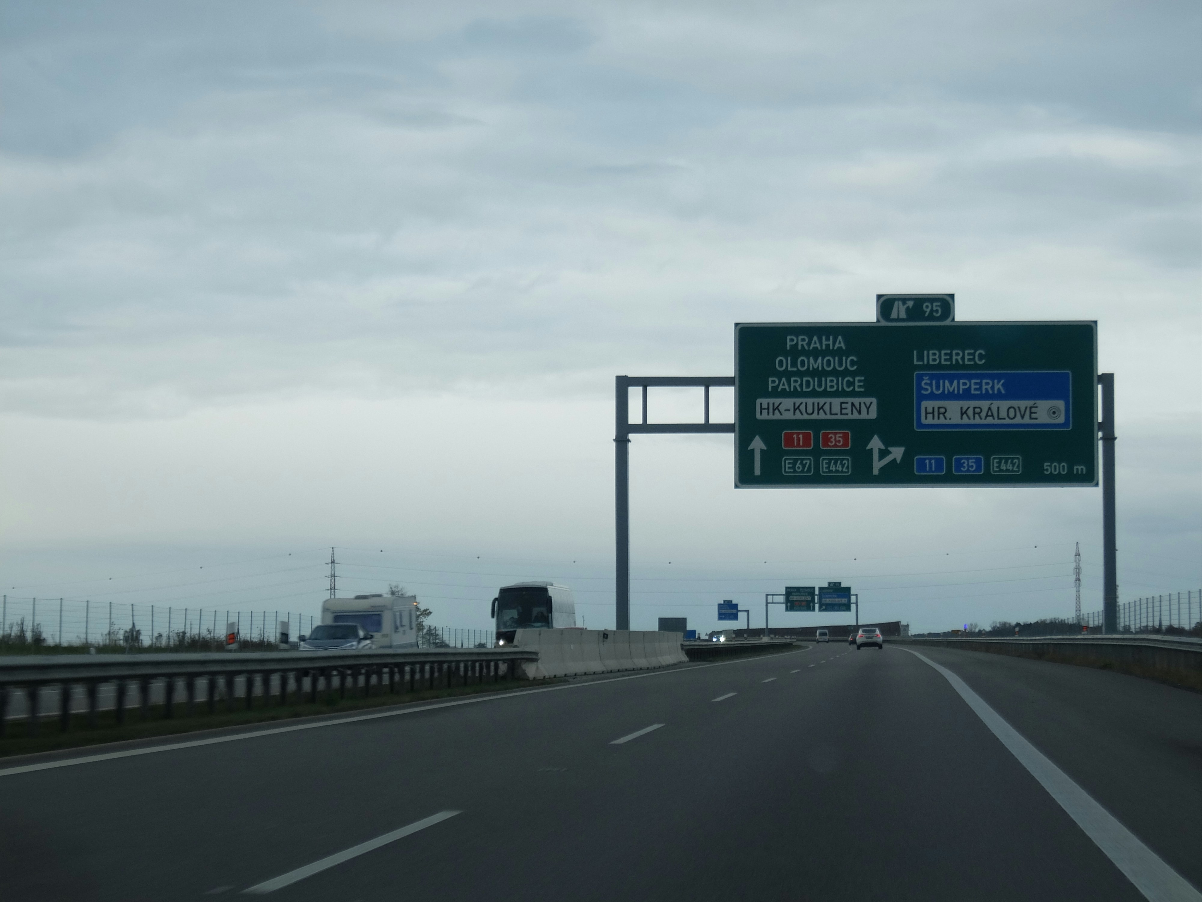 Highway scene featuring directional signs for various Czech cities under a cloudy sky. Traffic flows smoothly along the well-maintained road.