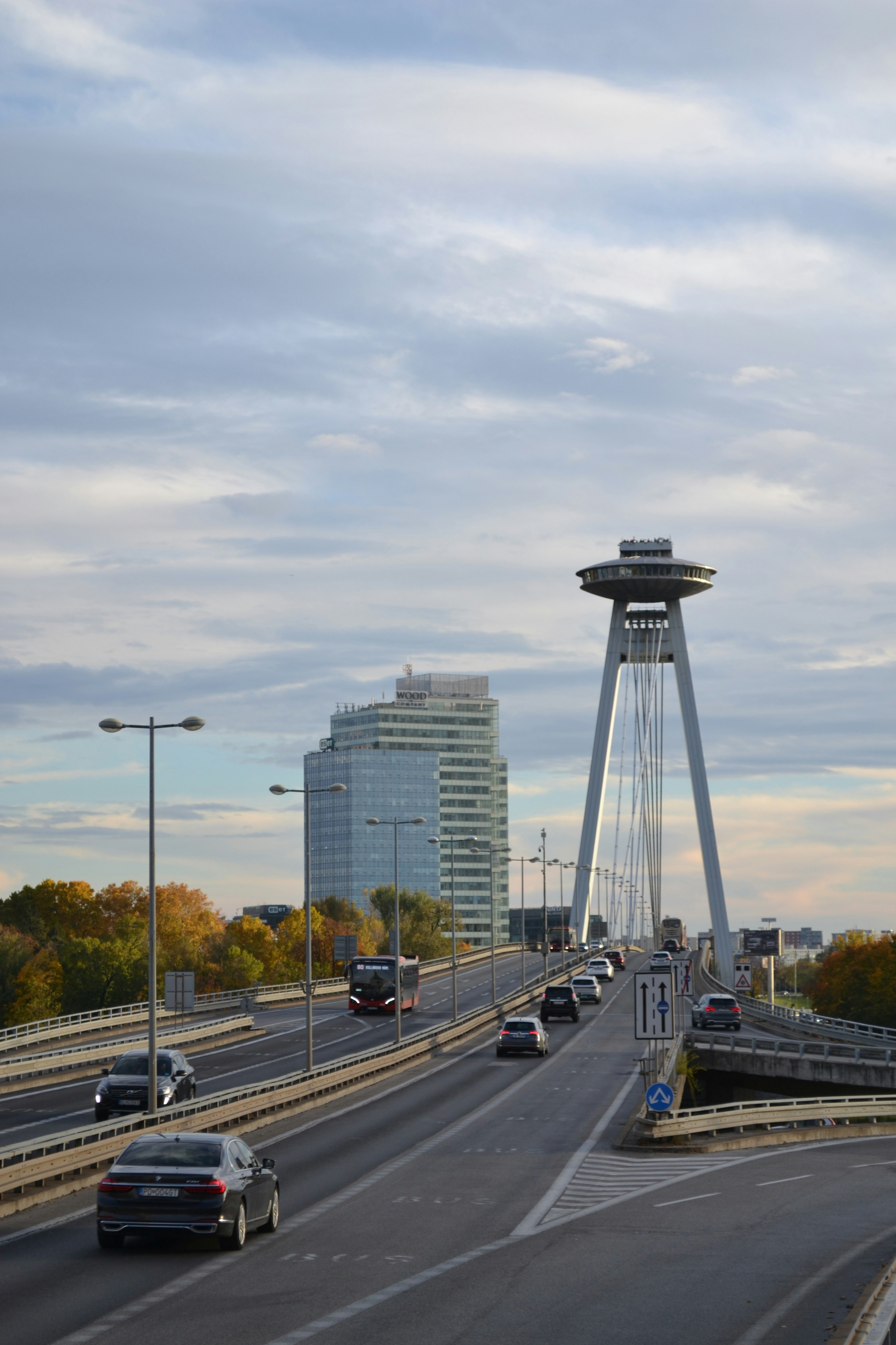 Suspension bridge with modern architecture spans a busy roadway, flanked by a contemporary building and autumn foliage.