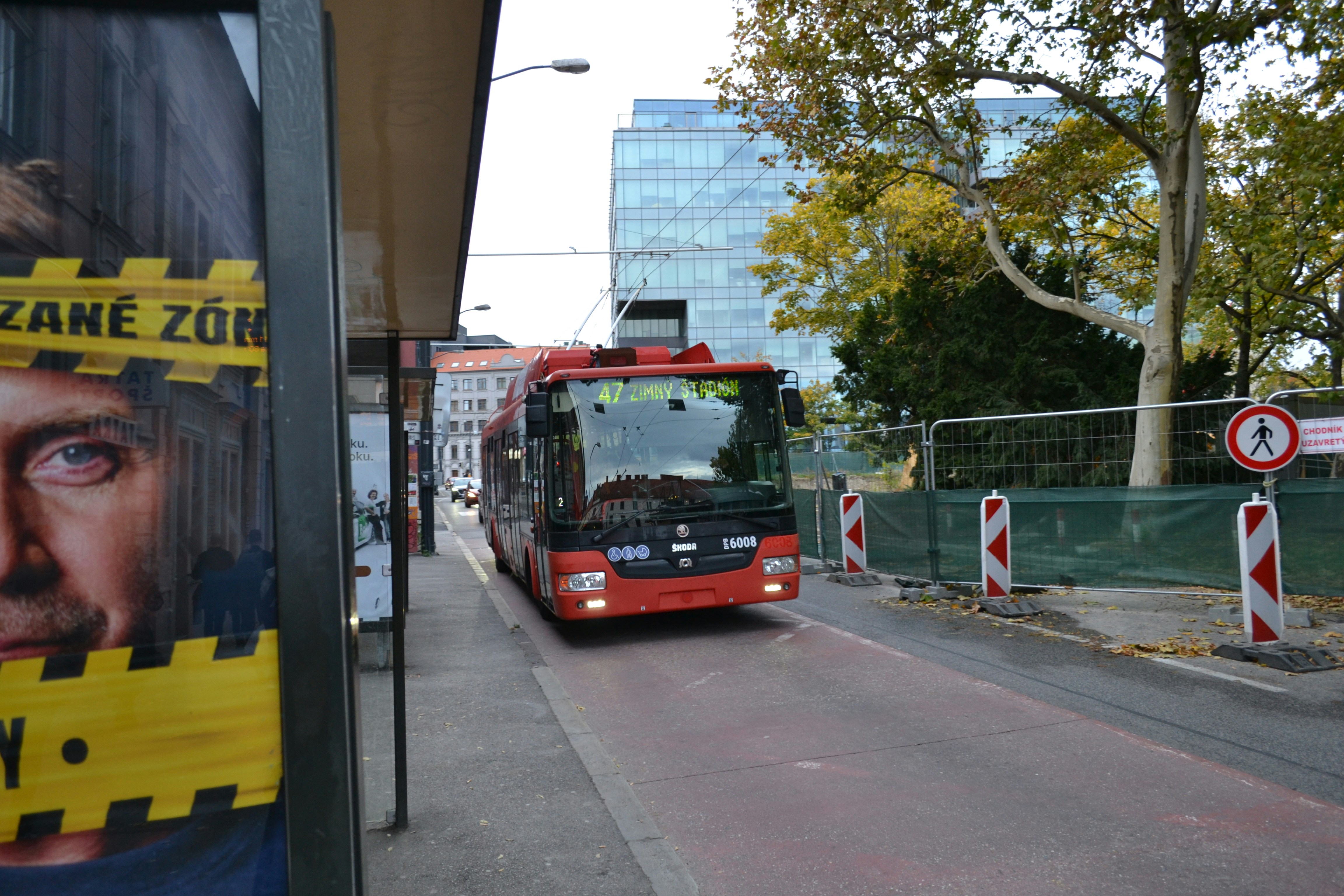 A red bus approaches a city stop, framed by construction barriers and an advertisement featuring a man's face. The scene captures the essence of urban commuting.
