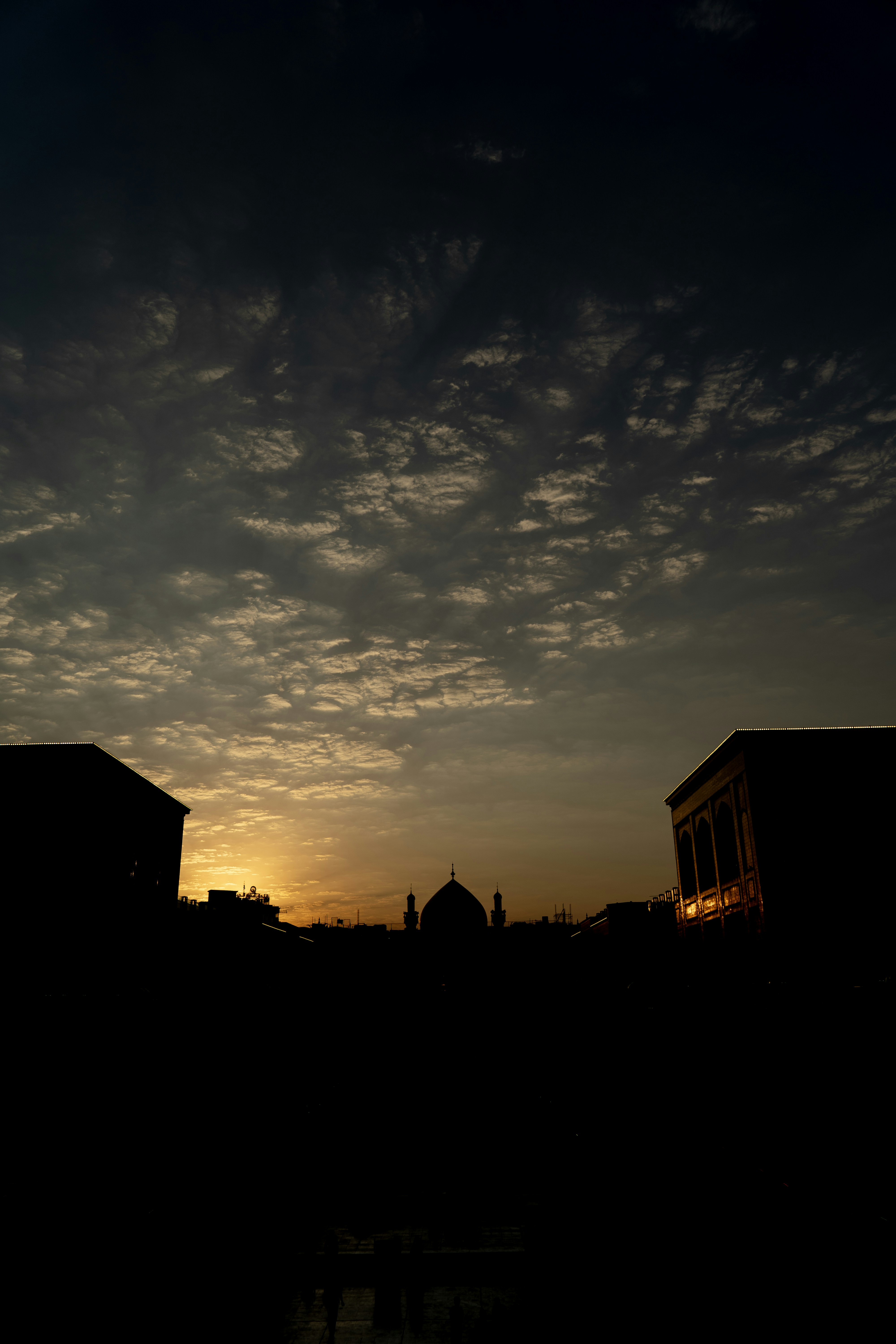 Silhouetted buildings frame a vibrant sunset sky filled with textured clouds, creating a dramatic evening atmosphere.