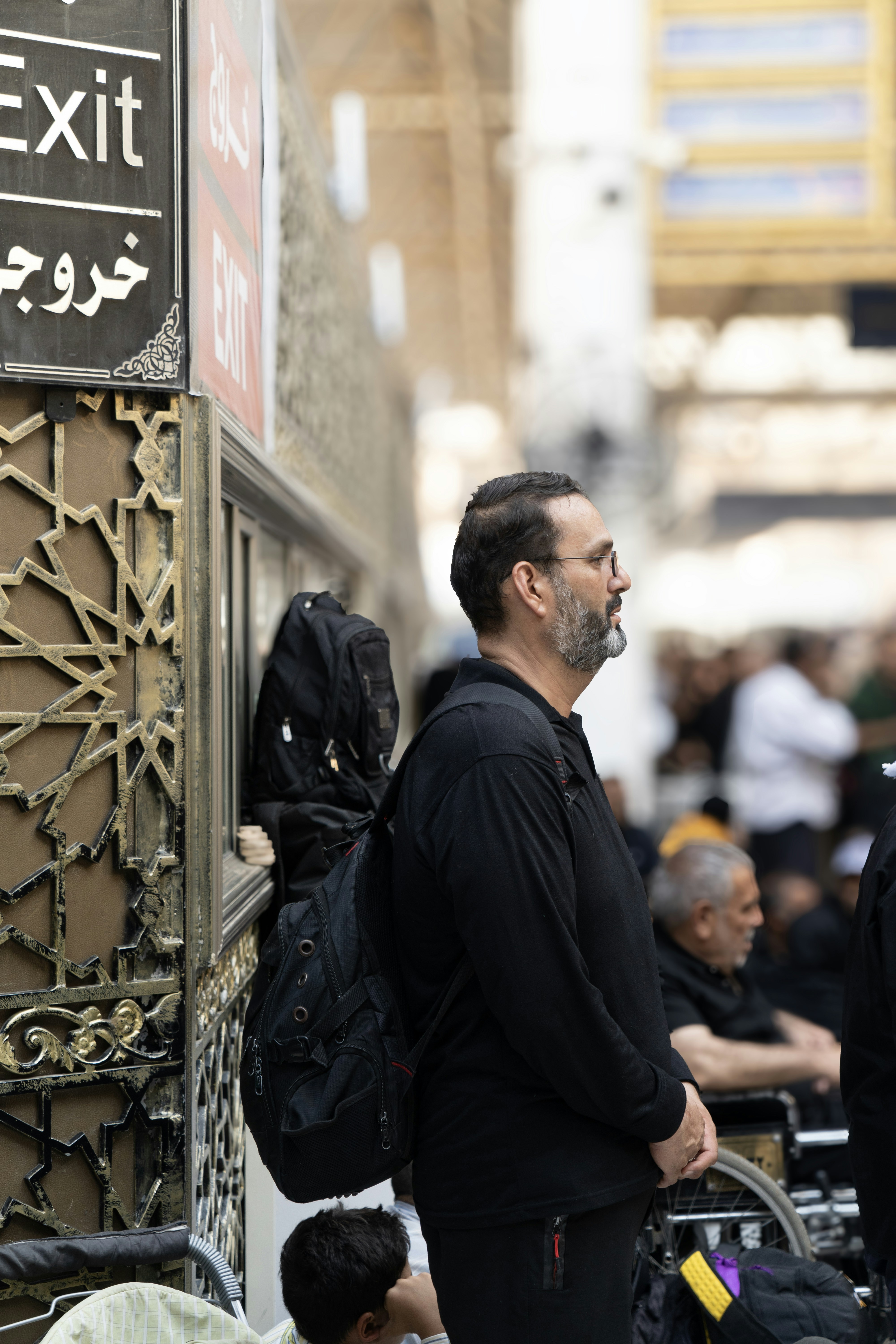 A man in black stands quietly near an ornate exit sign, surrounded by a bustling crowd. The scene captures a moment of reflection in a busy environment.