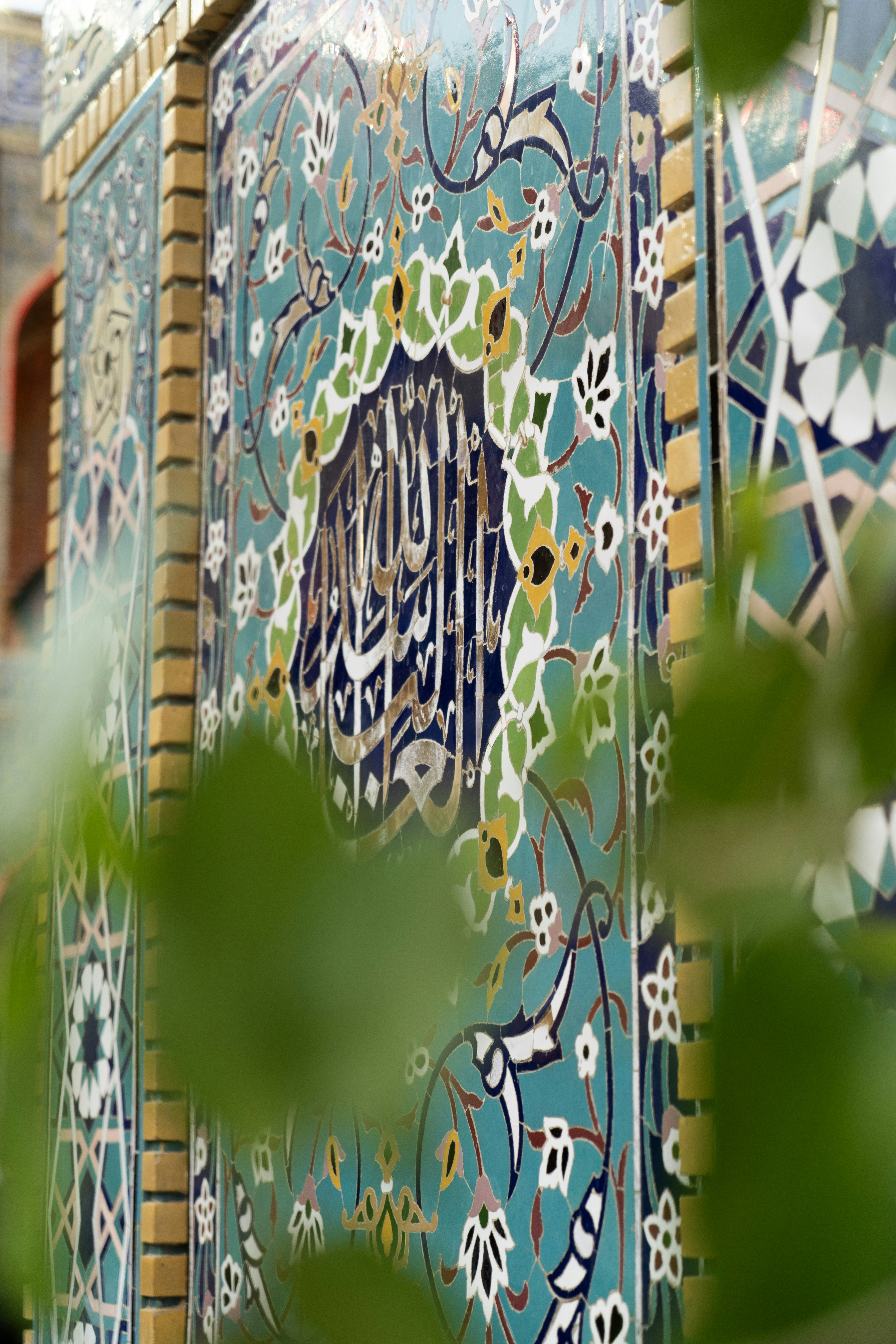 Vibrant tile work featuring intricate floral patterns and calligraphy, partially obscured by green foliage. The design reflects traditional craftsmanship and cultural significance.