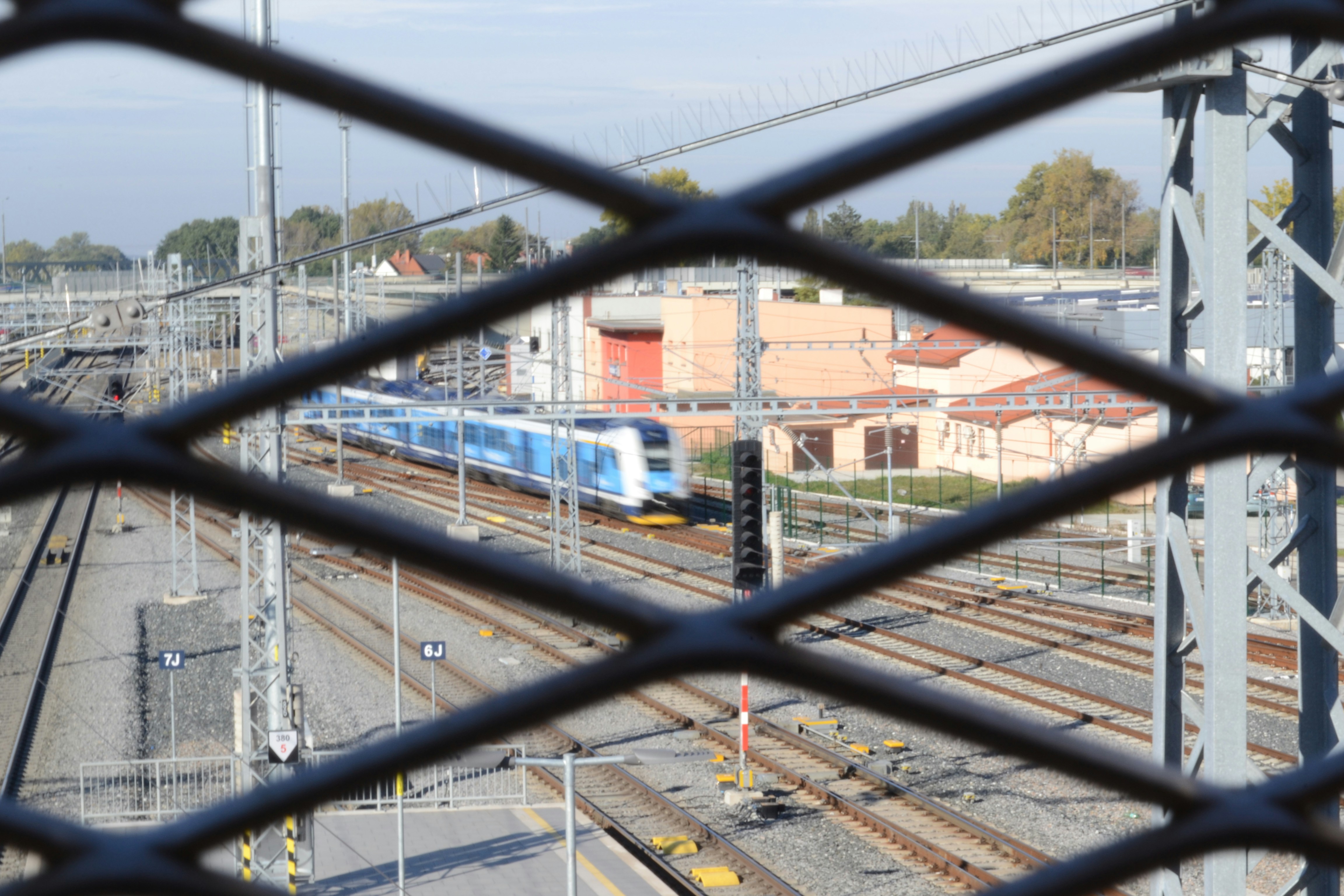 A blue train speeds past a railway crossing, viewed through a metal grate. The scene captures the dynamic energy of rail travel.