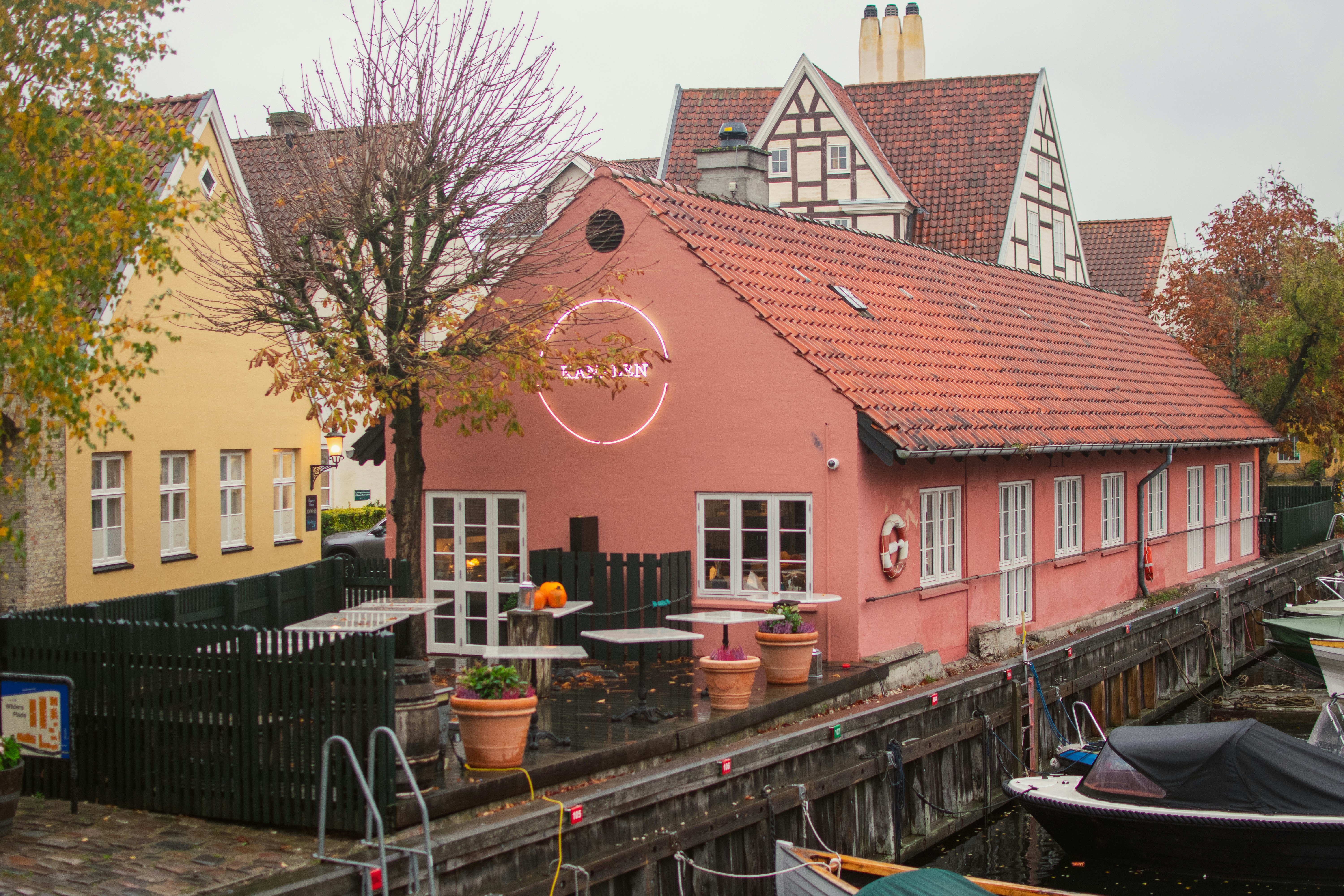 Charming pink waterfront building with outdoor seating and potted plants, nestled among autumn foliage. The inviting atmosphere is enhanced by the nearby boats and tranquil harbor.