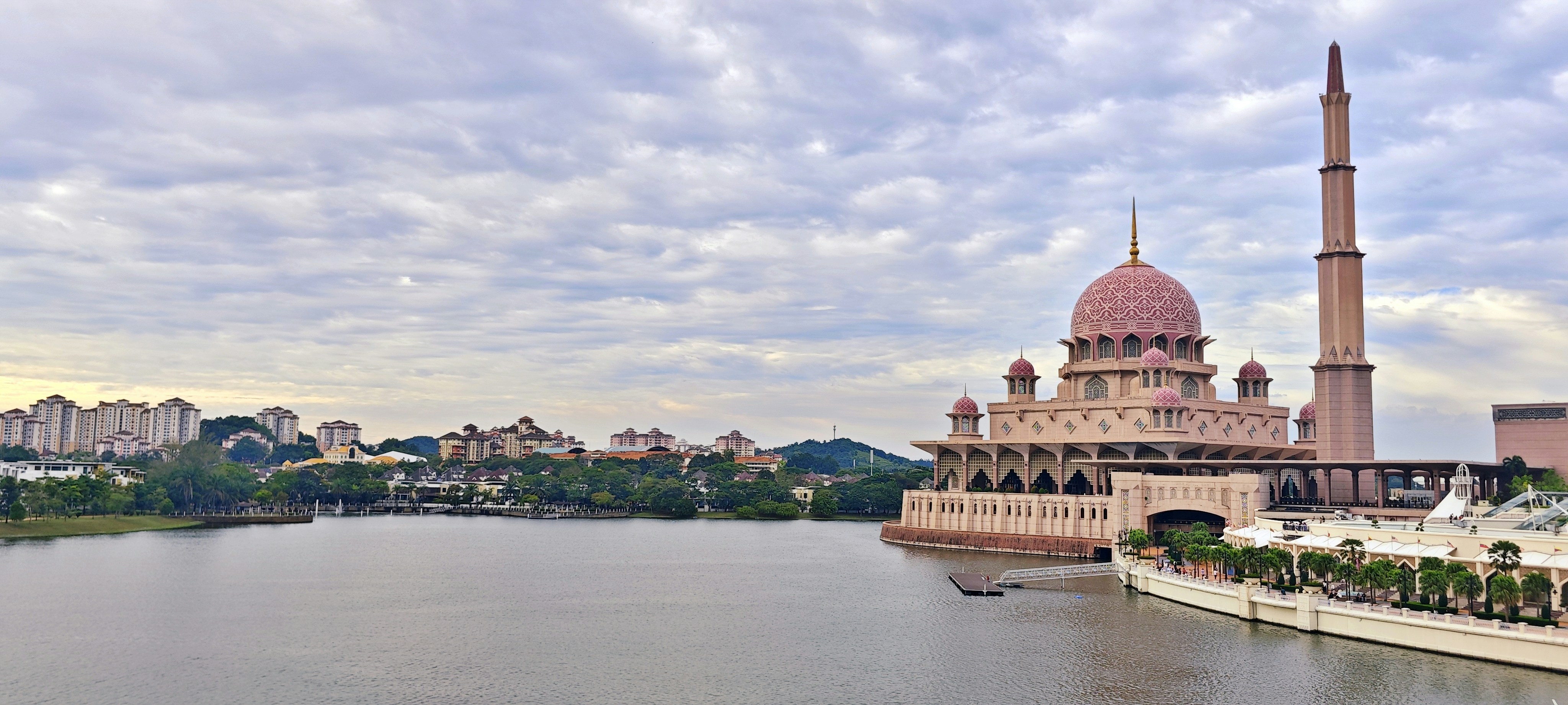 A majestic mosque with a pink dome and towering minaret beside a calm lake, framed by a backdrop of modern buildings and a cloudy sky.