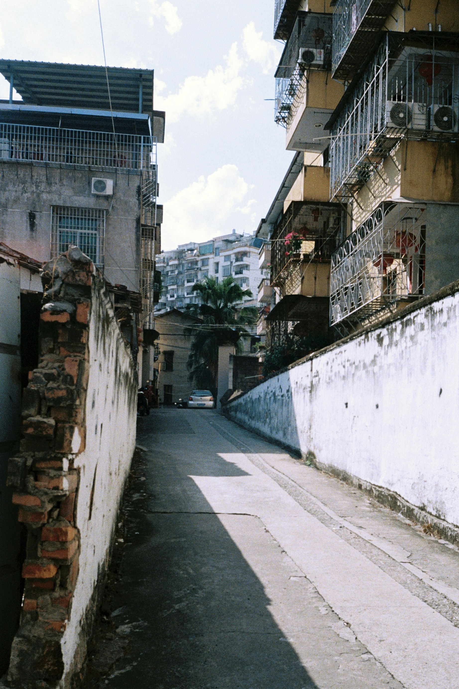 Narrow alleyway flanked by aged brick walls and modern apartment buildings, leading to a distant street with a glimpse of palm trees. 