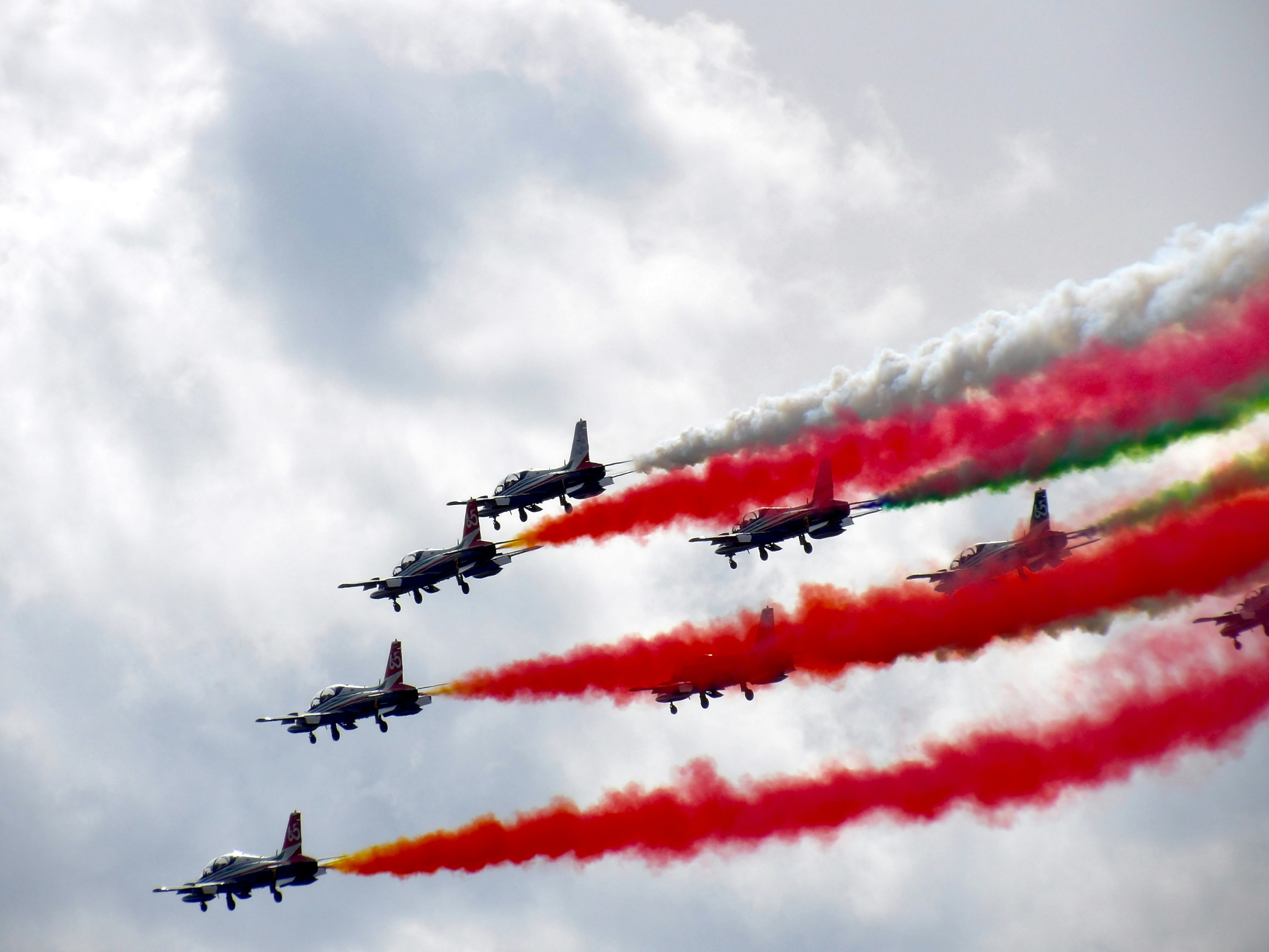 Formation of aircraft releasing vibrant smoke trails in red, green, and yellow against a cloudy backdrop.