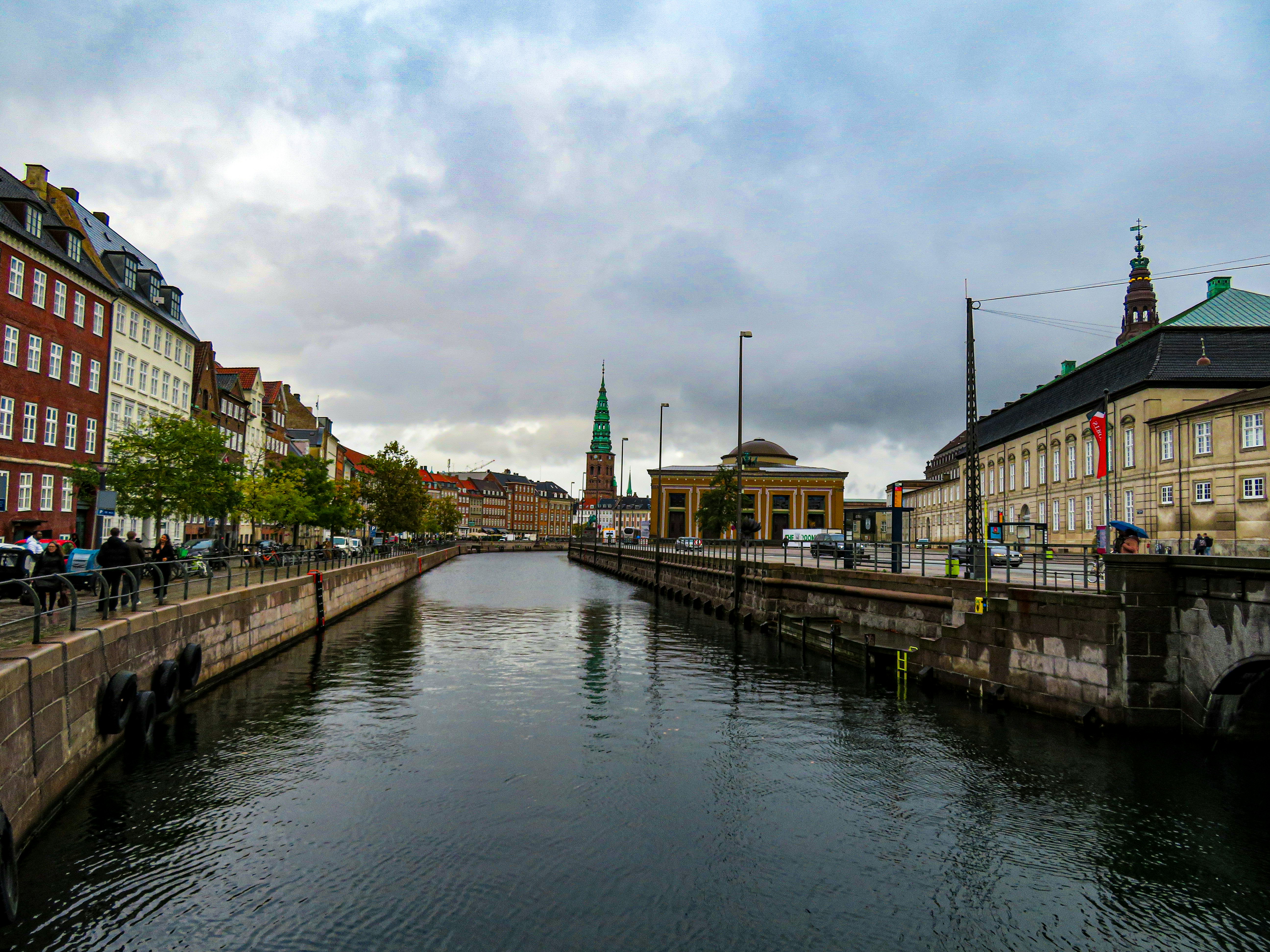 Canal in Copenhagen lined with historic buildings and a prominent church spire in the background. The calm water reflects the surrounding architecture.
