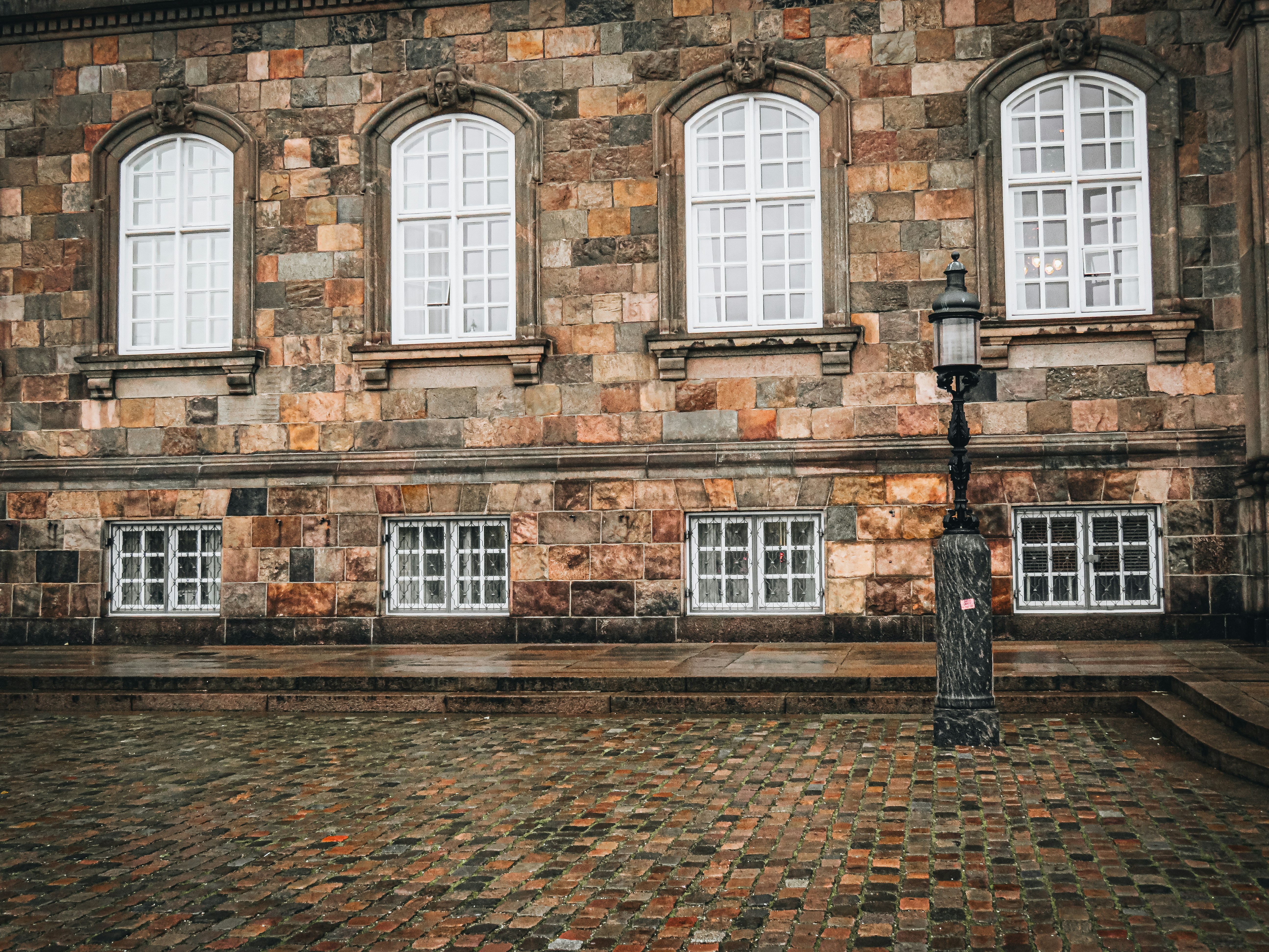 Historic stone building facade featuring arched windows and a vintage lamp post, set against a cobblestone path.