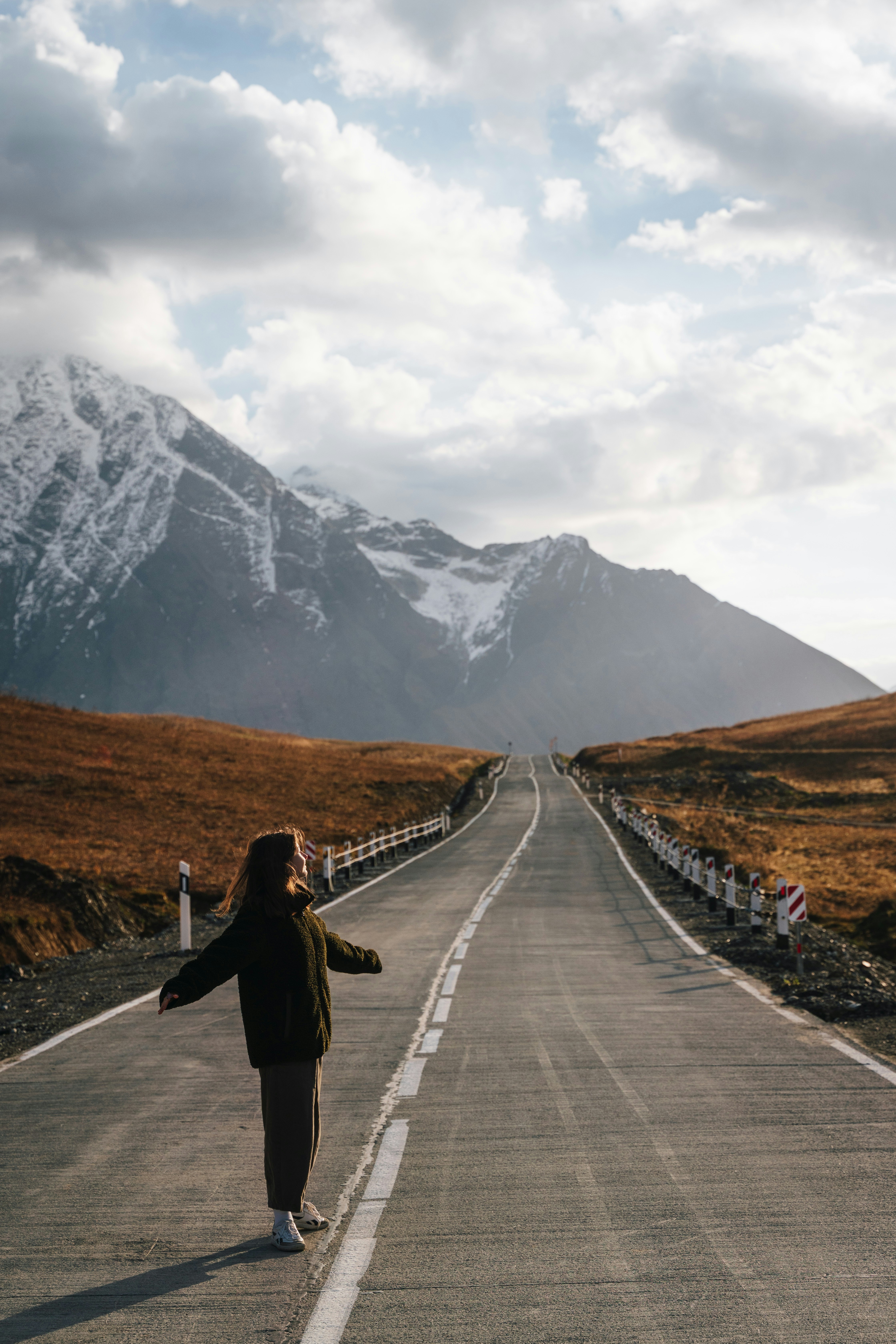 A woman stands with arms outstretched on an empty road, framed by majestic mountains and a vast sky. The scene captures a moment of freedom and connection with nature.