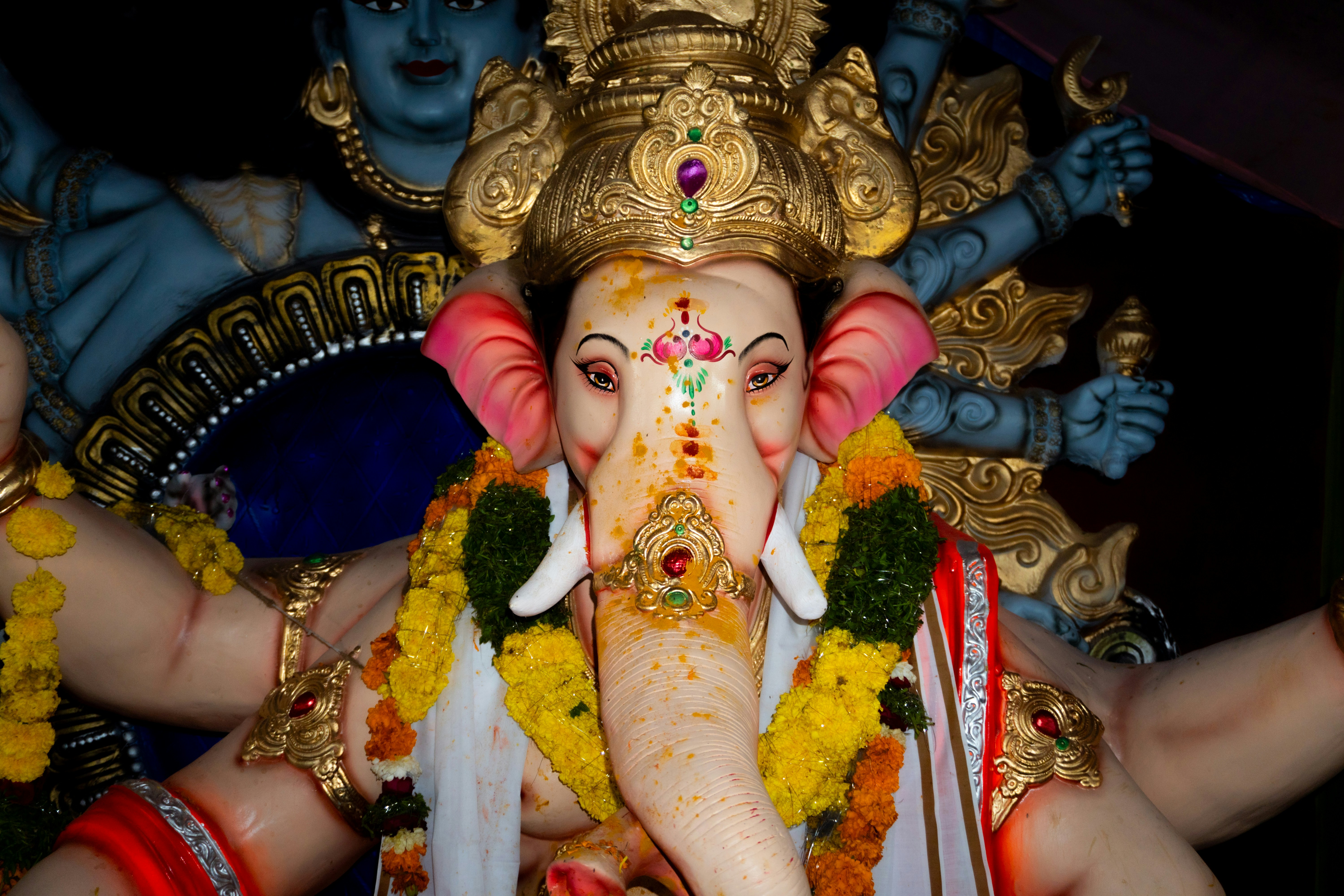 Colorful idol of Lord Ganesha adorned with flowers and intricate jewelry, set against a backdrop of deities. The craftsmanship highlights cultural devotion.