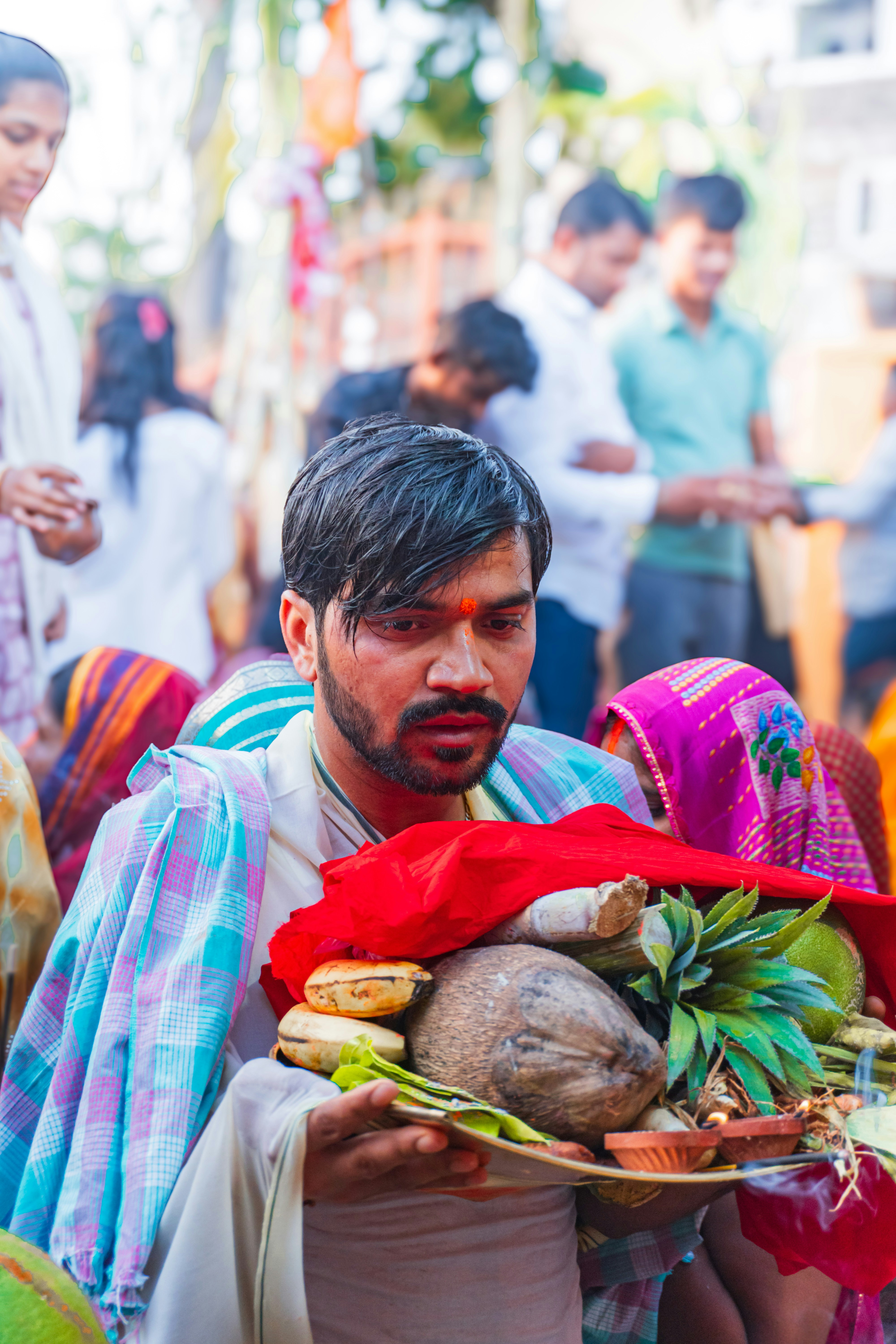 Man in traditional attire carries a ceremonial offering of fruits and a coconut during a festive celebration, surrounded by participants in vibrant clothing.