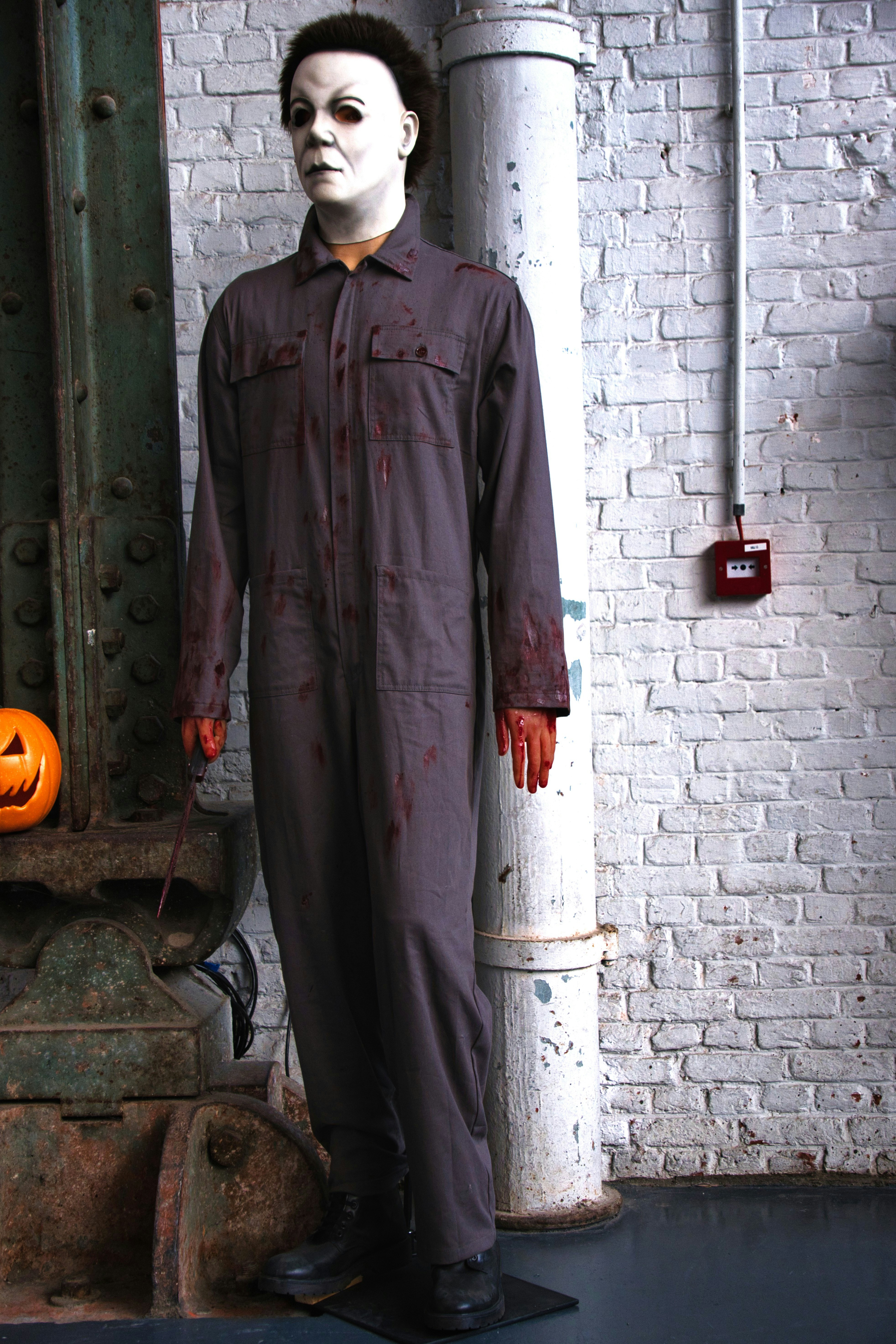 A life-sized mannequin dressed in a blood-stained jumpsuit, embodying a classic horror character, stands ominously next to a carved pumpkin in an industrial setting.