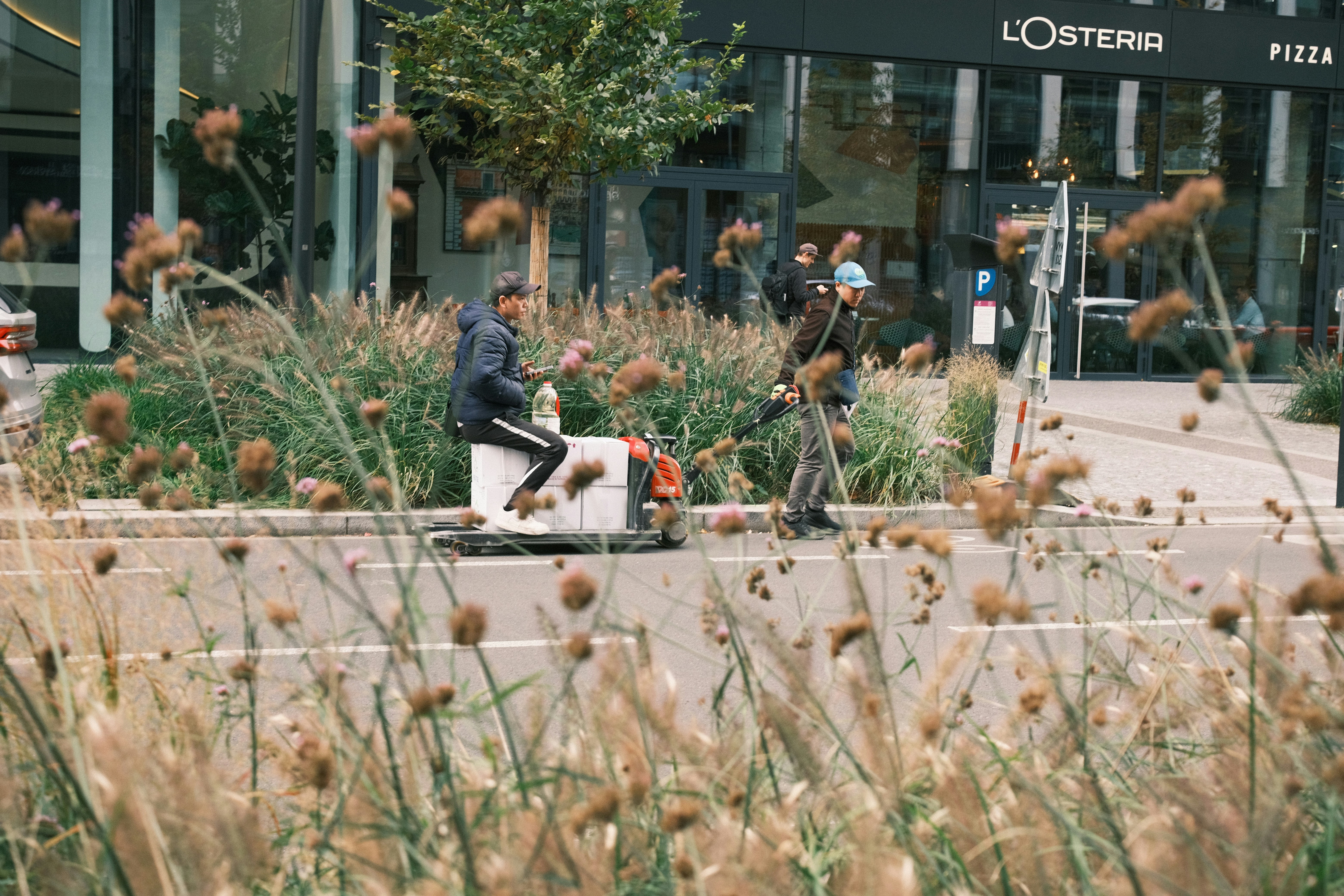 Two individuals navigate a lively urban scene, framed by lush grass and modern architecture. The interaction captures the essence of city life.