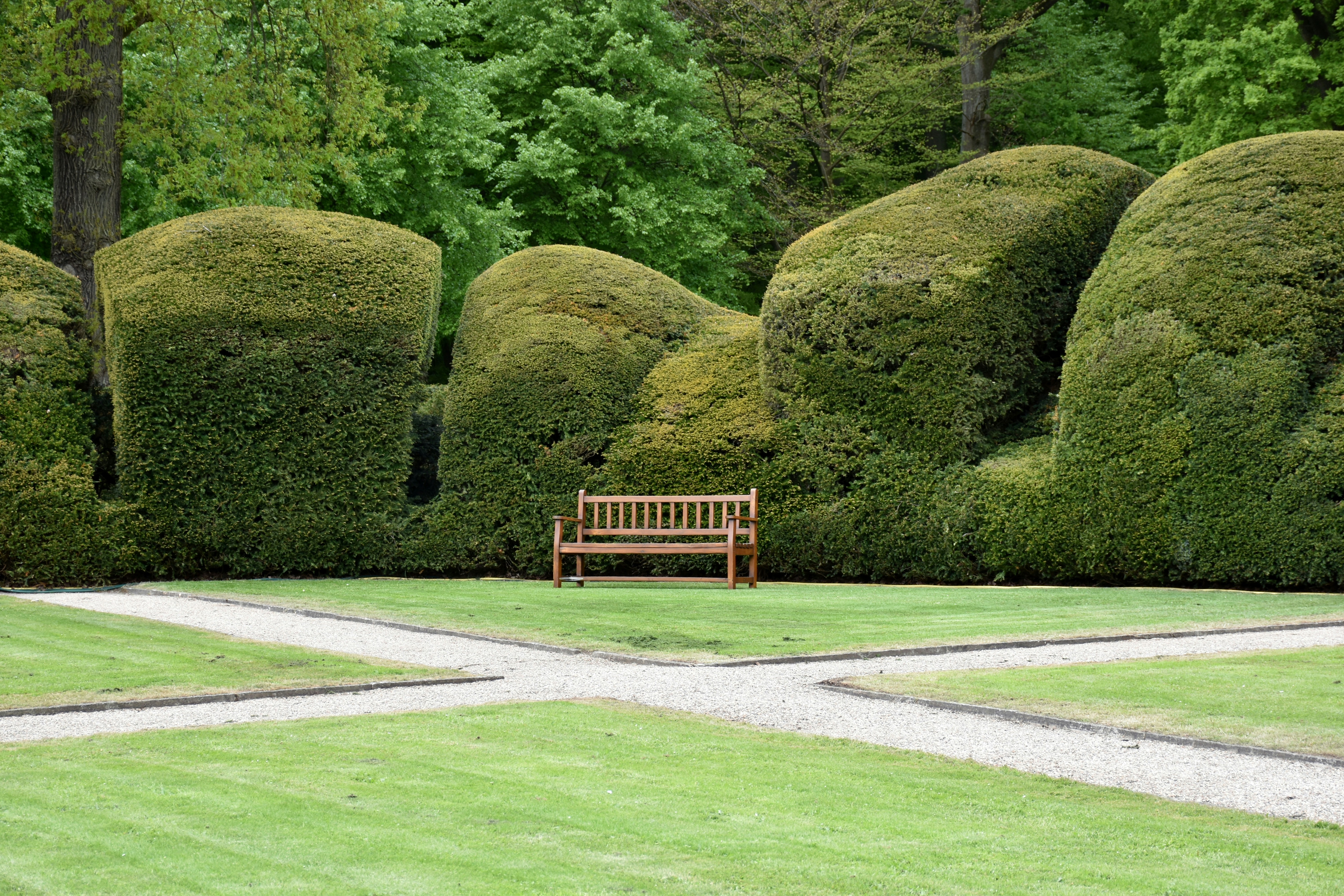 A wooden bench nestled within meticulously trimmed hedges, surrounded by intersecting gravel paths in a tranquil garden setting.