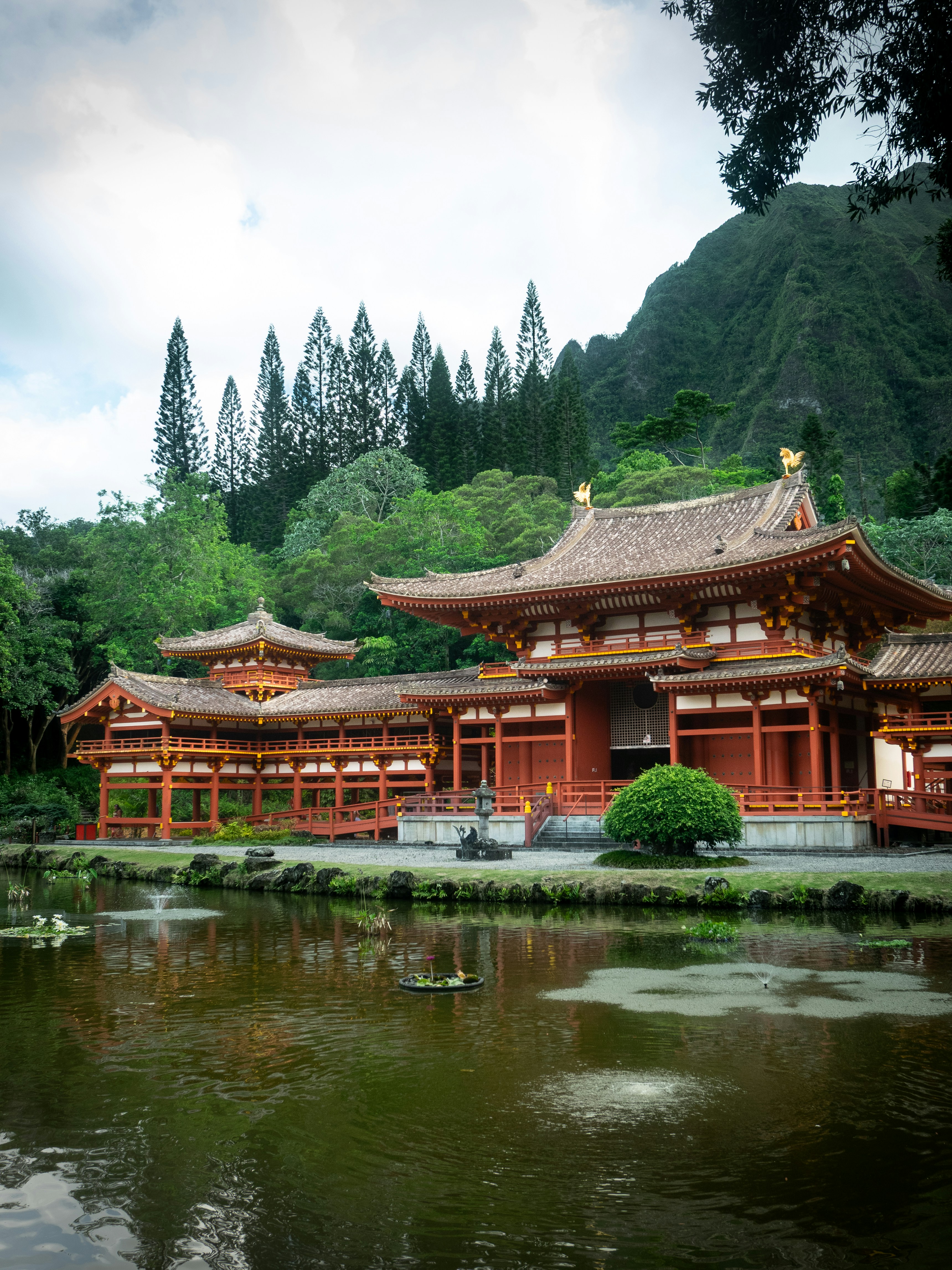 Traditional Japanese temple nestled beside a tranquil pond, surrounded by lush greenery and towering trees.