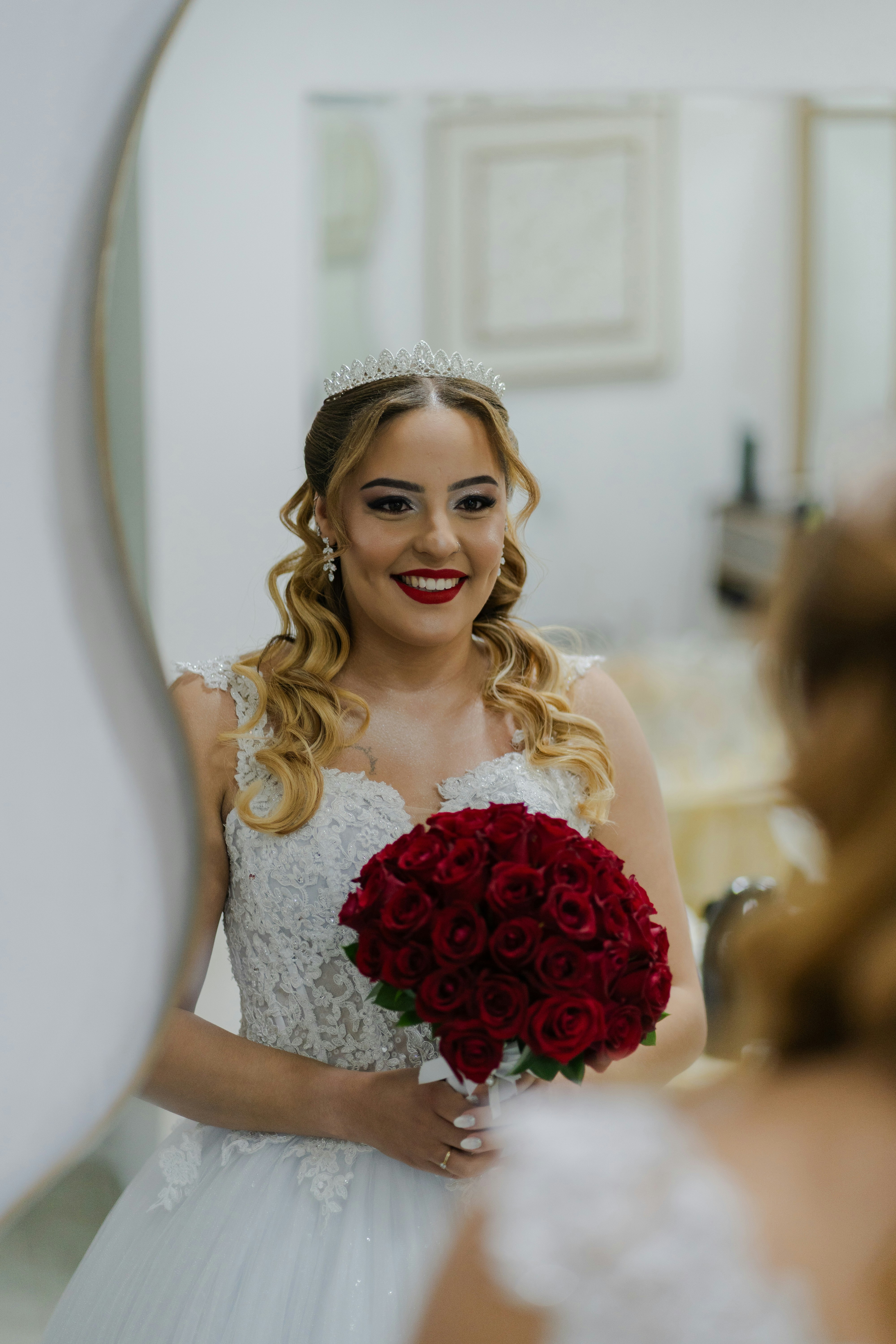 Bride admiring herself in a mirror while holding a bouquet of red roses, radiating happiness and elegance.