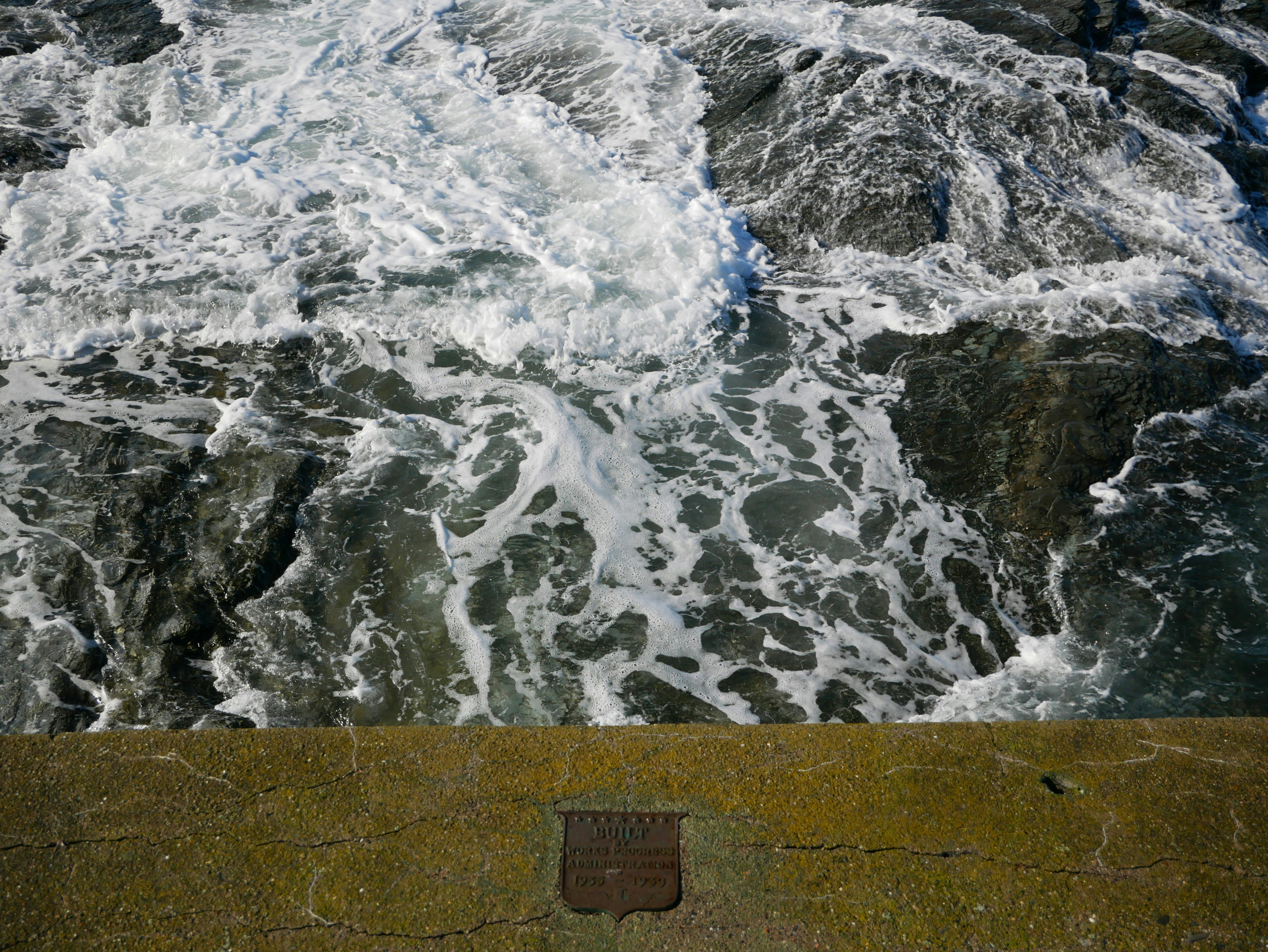 Foamy waves crash against a rugged shoreline, highlighting the dynamic interplay between water and rock. A plaque rests on the edge, hinting at the area's significance.