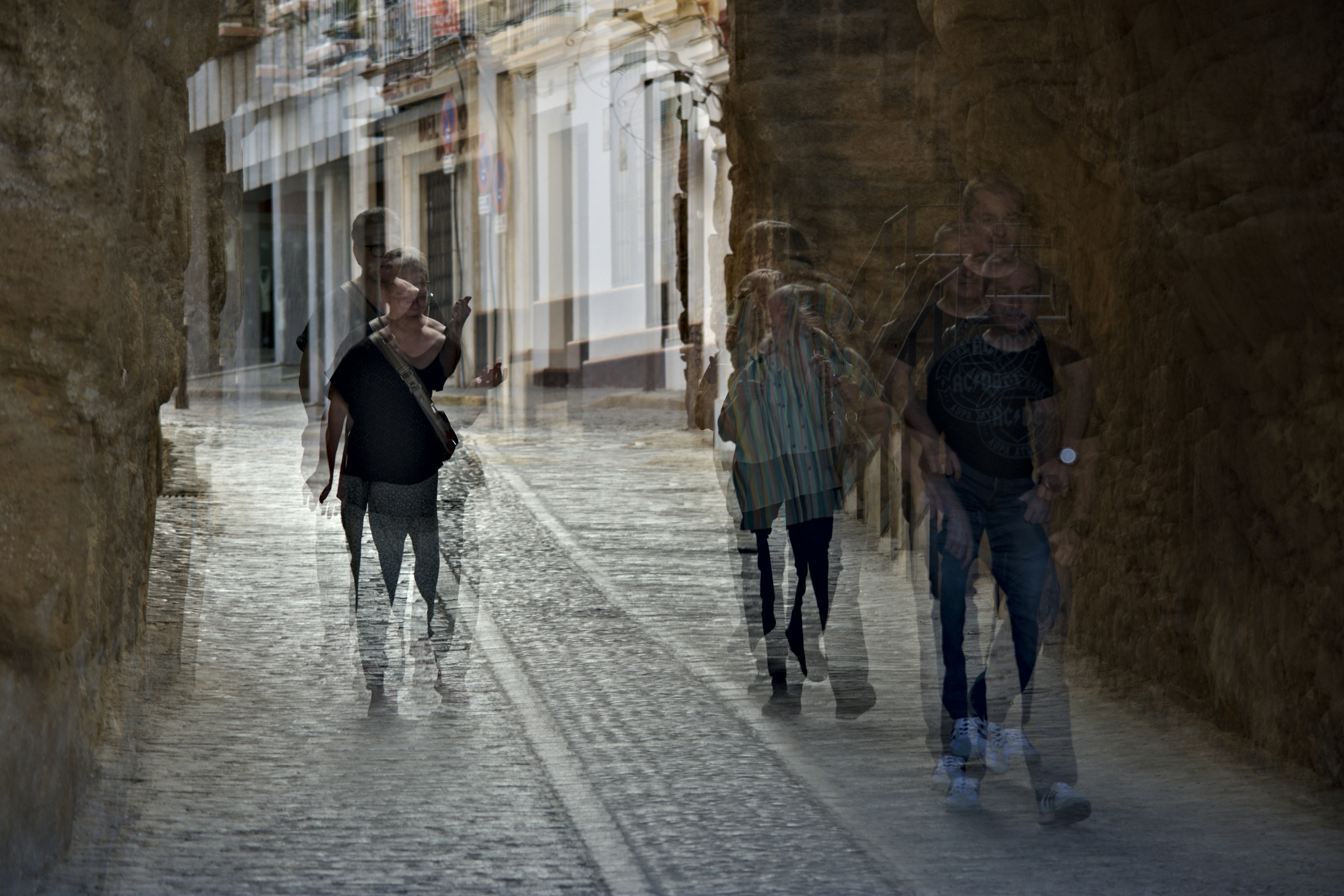 Multiple figures walk through a sunlit cobblestone alley, captured with a motion blur effect that conveys a sense of movement and life.