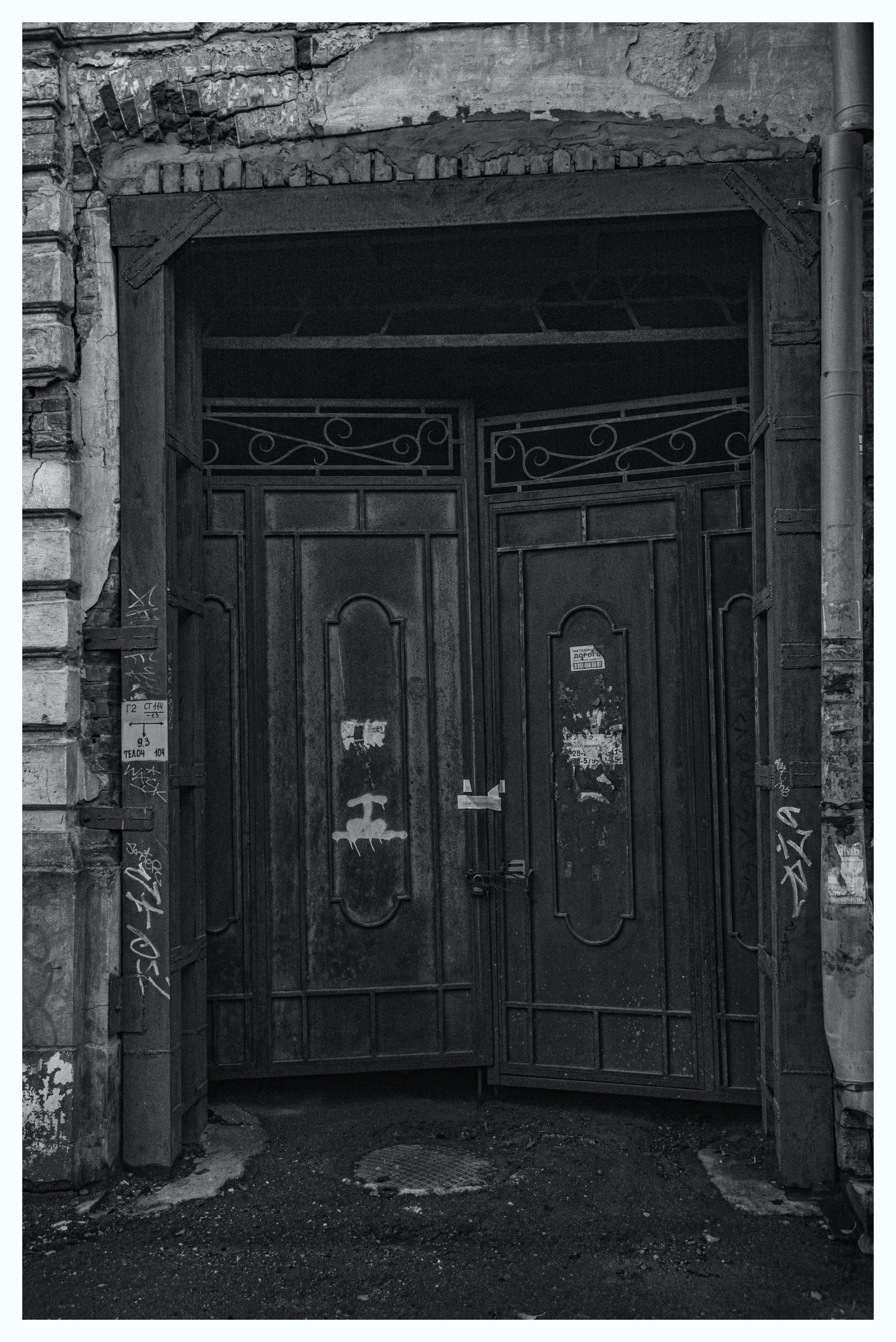Weathered double doors adorned with graffiti, set against a crumbling wall, evoke a sense of forgotten history. The stark black and white tones enhance the mood of desolation.