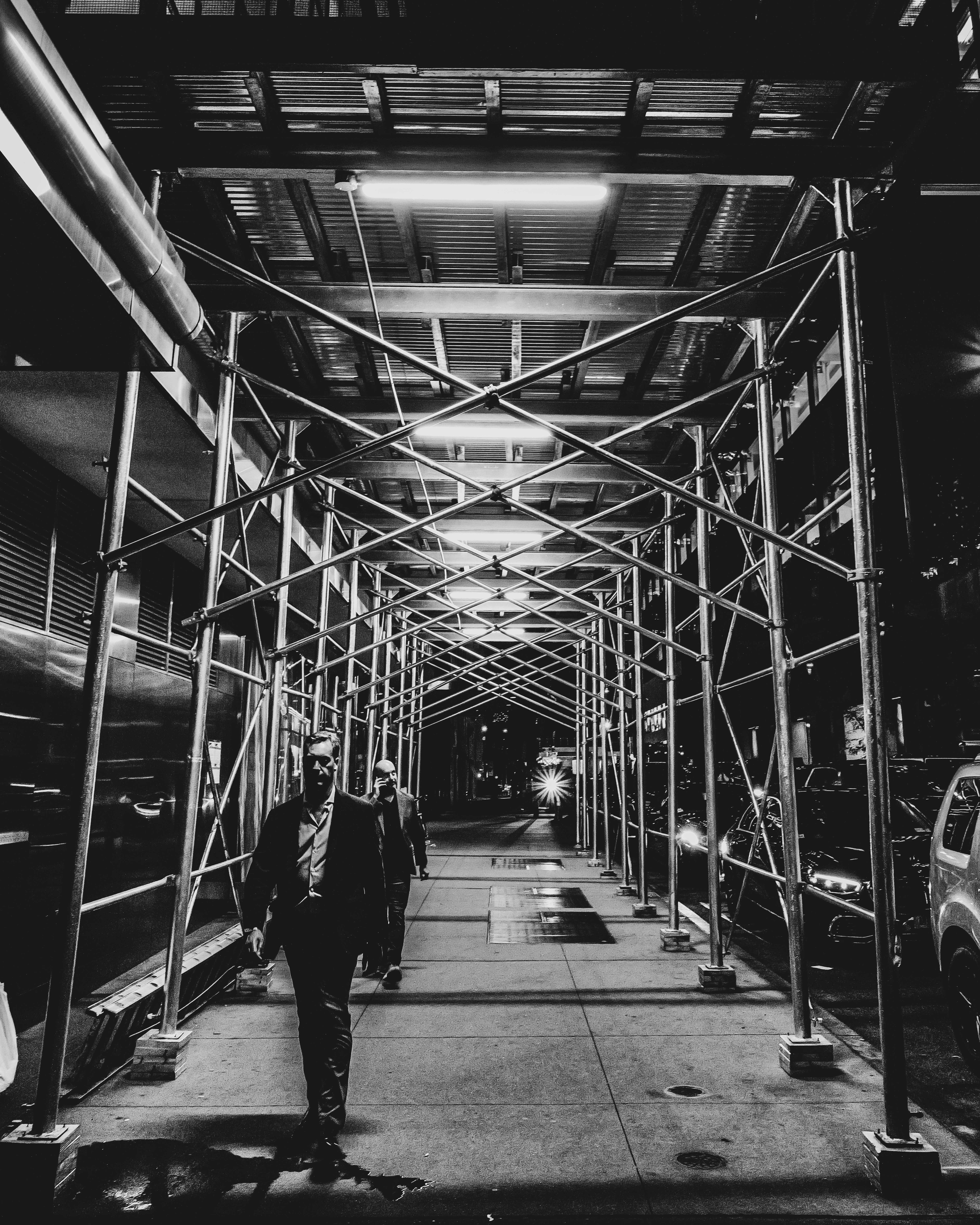 Men walking through a scaffolded urban corridor illuminated by overhead lights, creating a striking interplay of light and shadow.