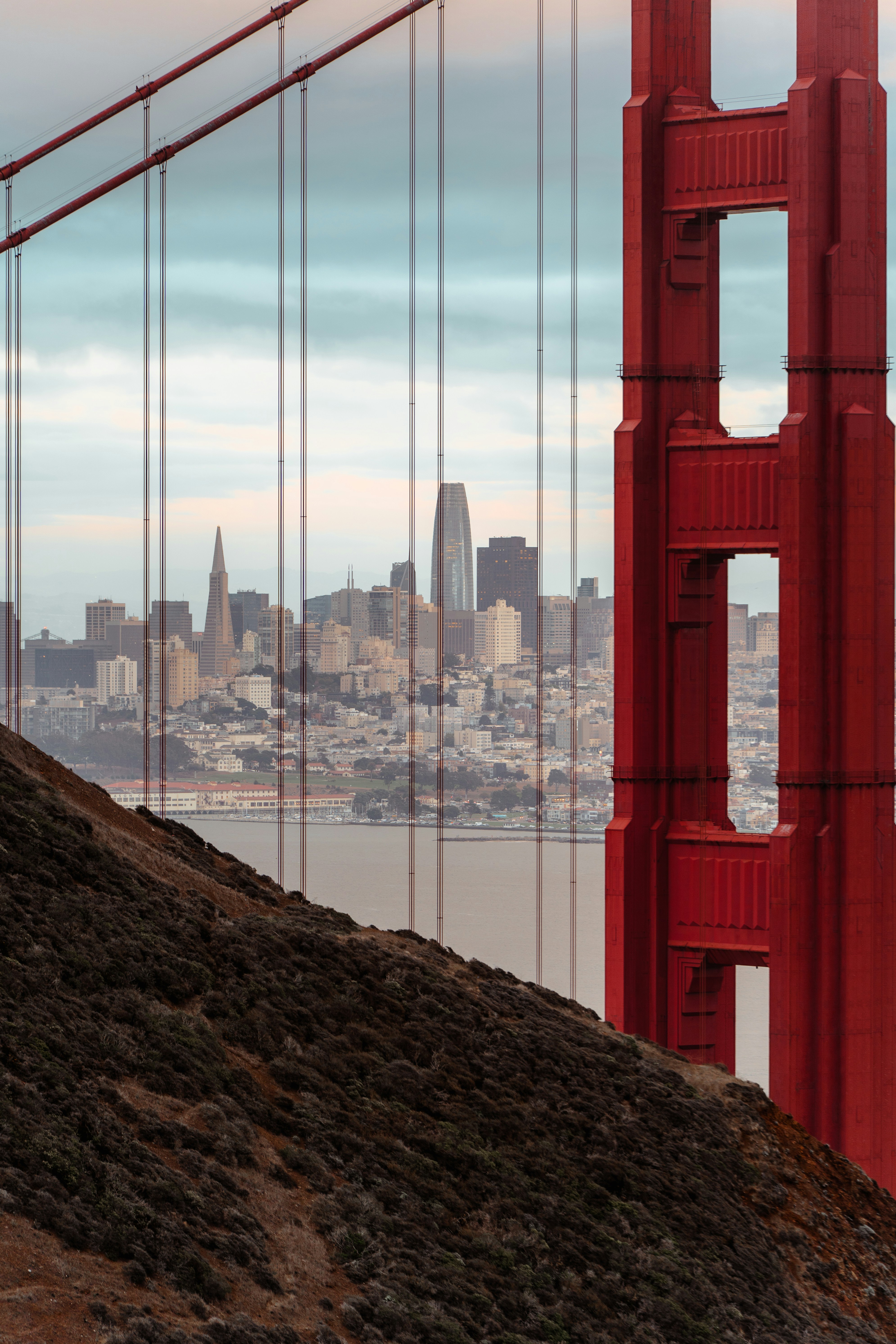 Ein detailreicher Blick durch die markanten Stahlstreben der Golden Gate Bridge auf die Skyline von San Francisco. Das ikonische Rot des Brückenpfeilers trifft auf die kühlen Pastelltöne des Morgens – ein Moment zwischen Architektur, Natur und urbaner Ruhe.