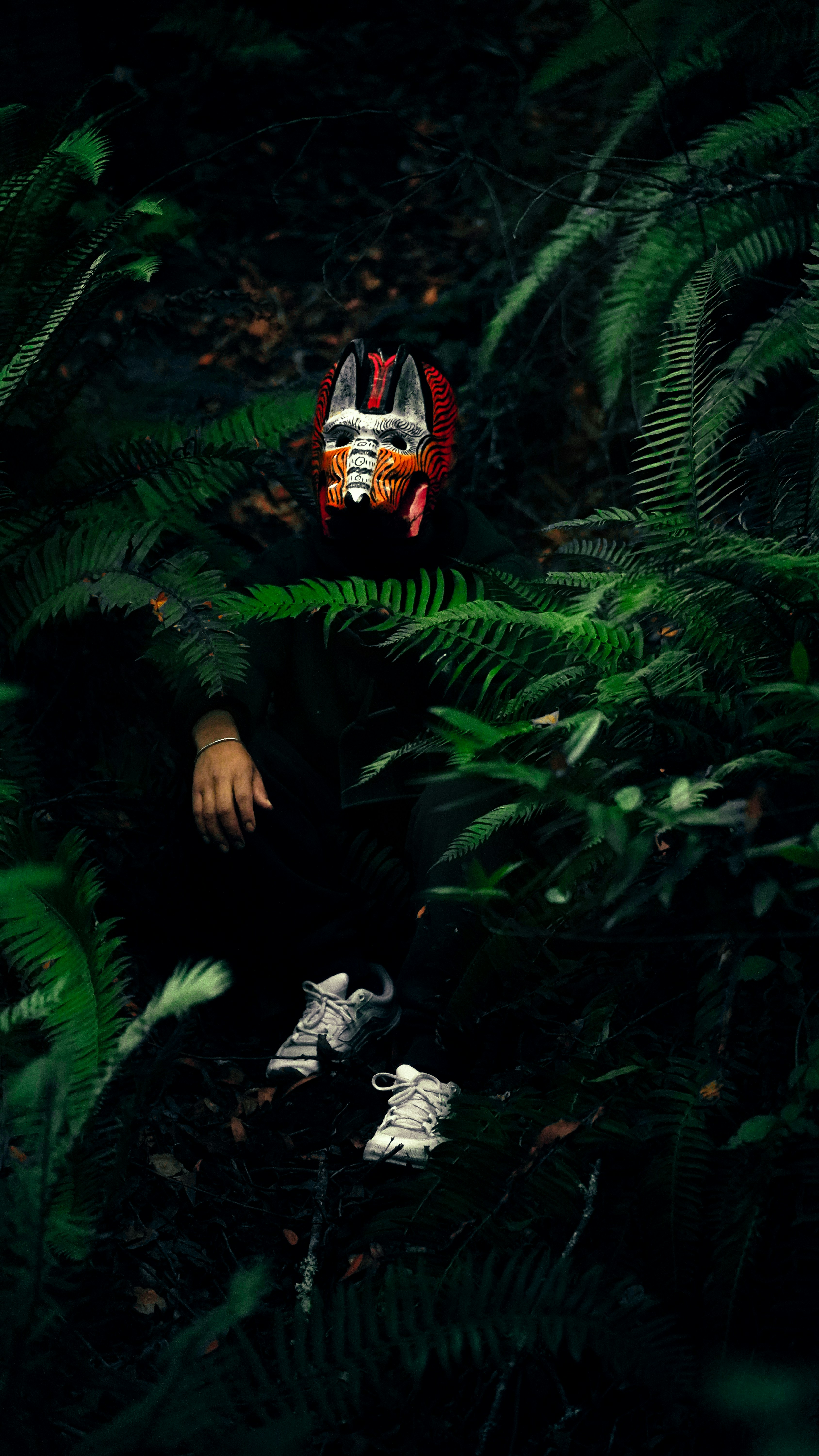 Person in a mask stands in a dark, leafy forest.
