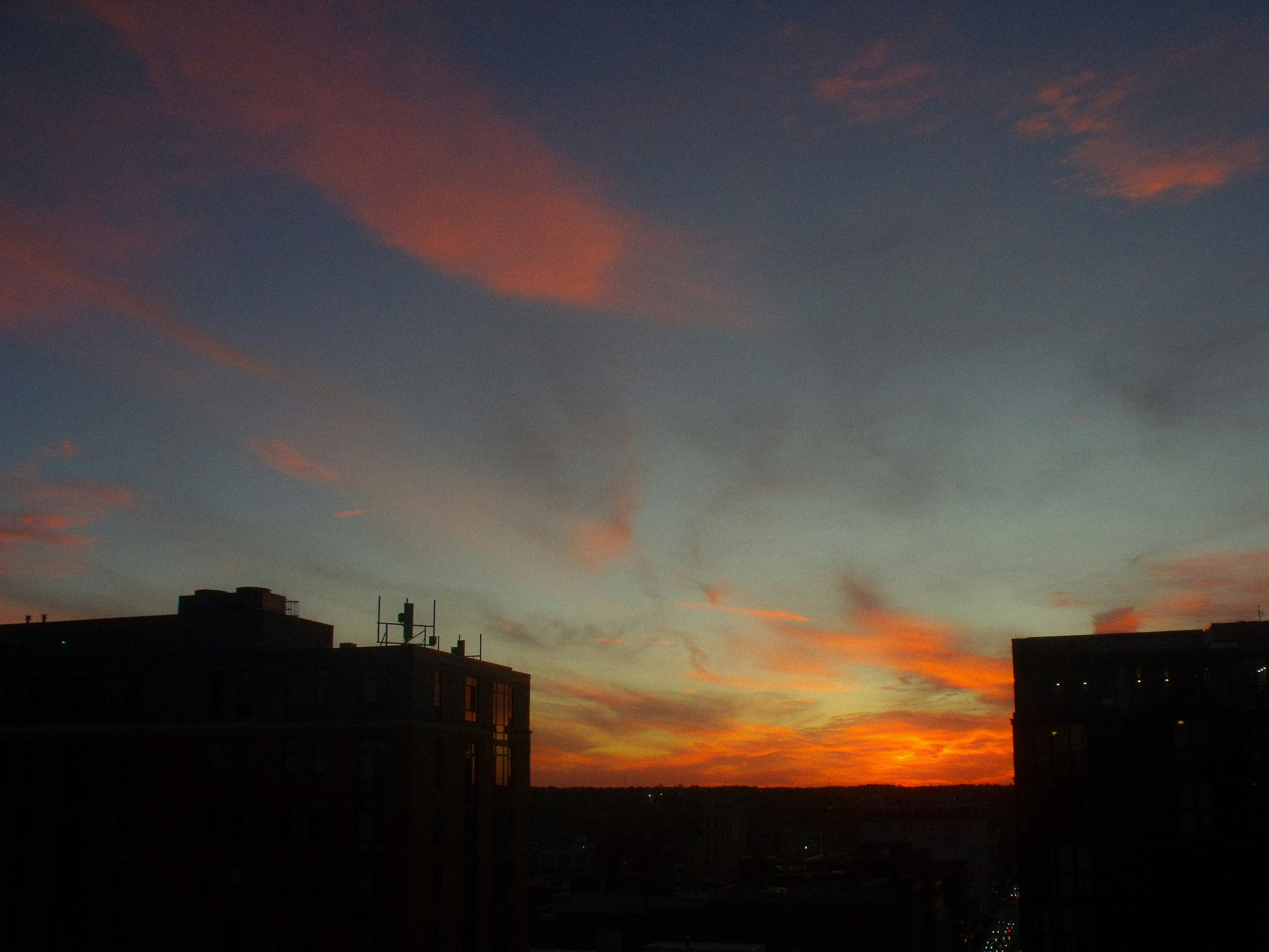 Vibrant sunset colors blend across the sky, silhouetting urban buildings against the horizon. The scene captures the transition from day to night.