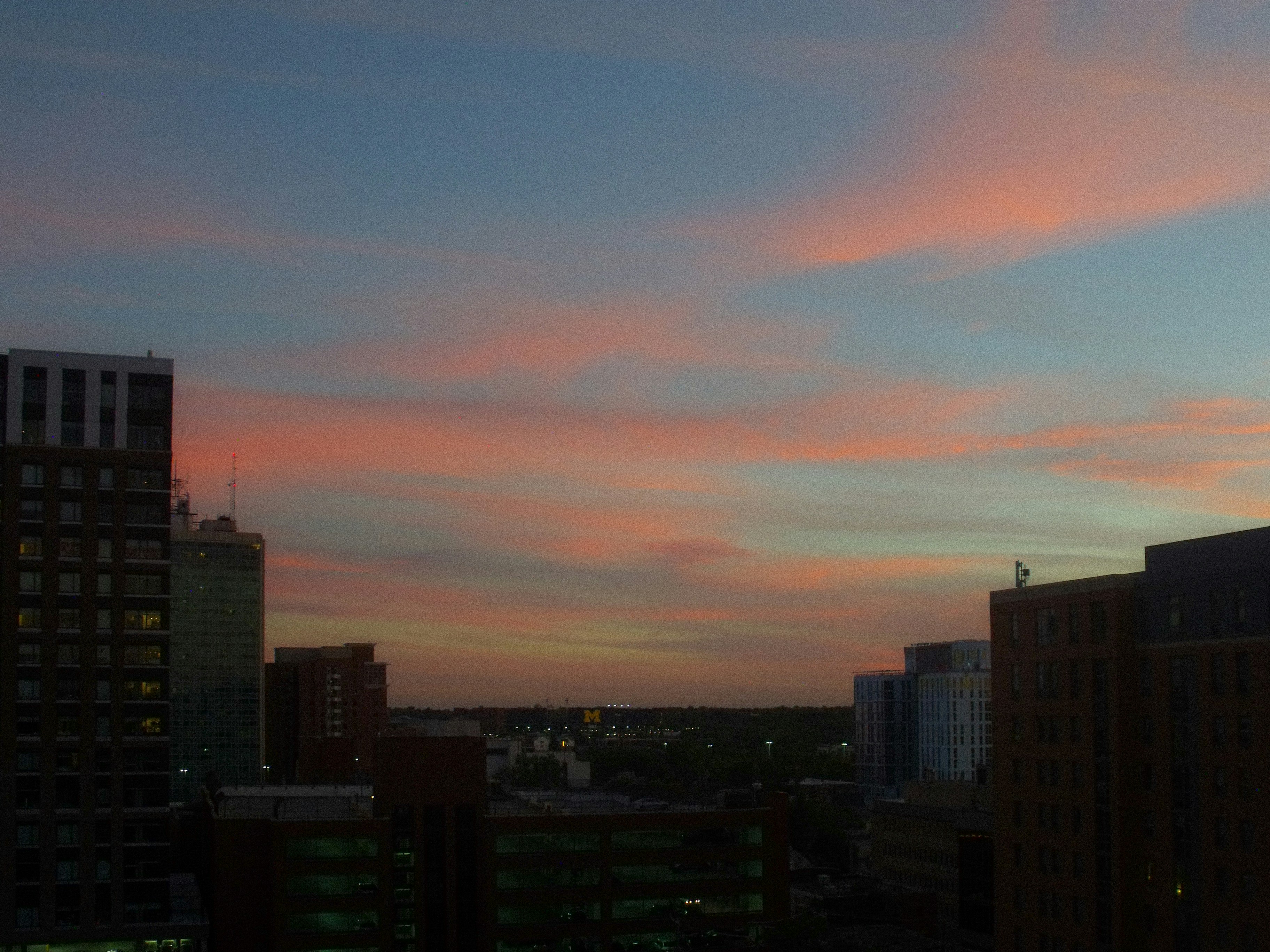 Soft pastel hues paint the evening sky above a city skyline, blending urban structures with nature's palette. The scene captures the transition from day to night.
