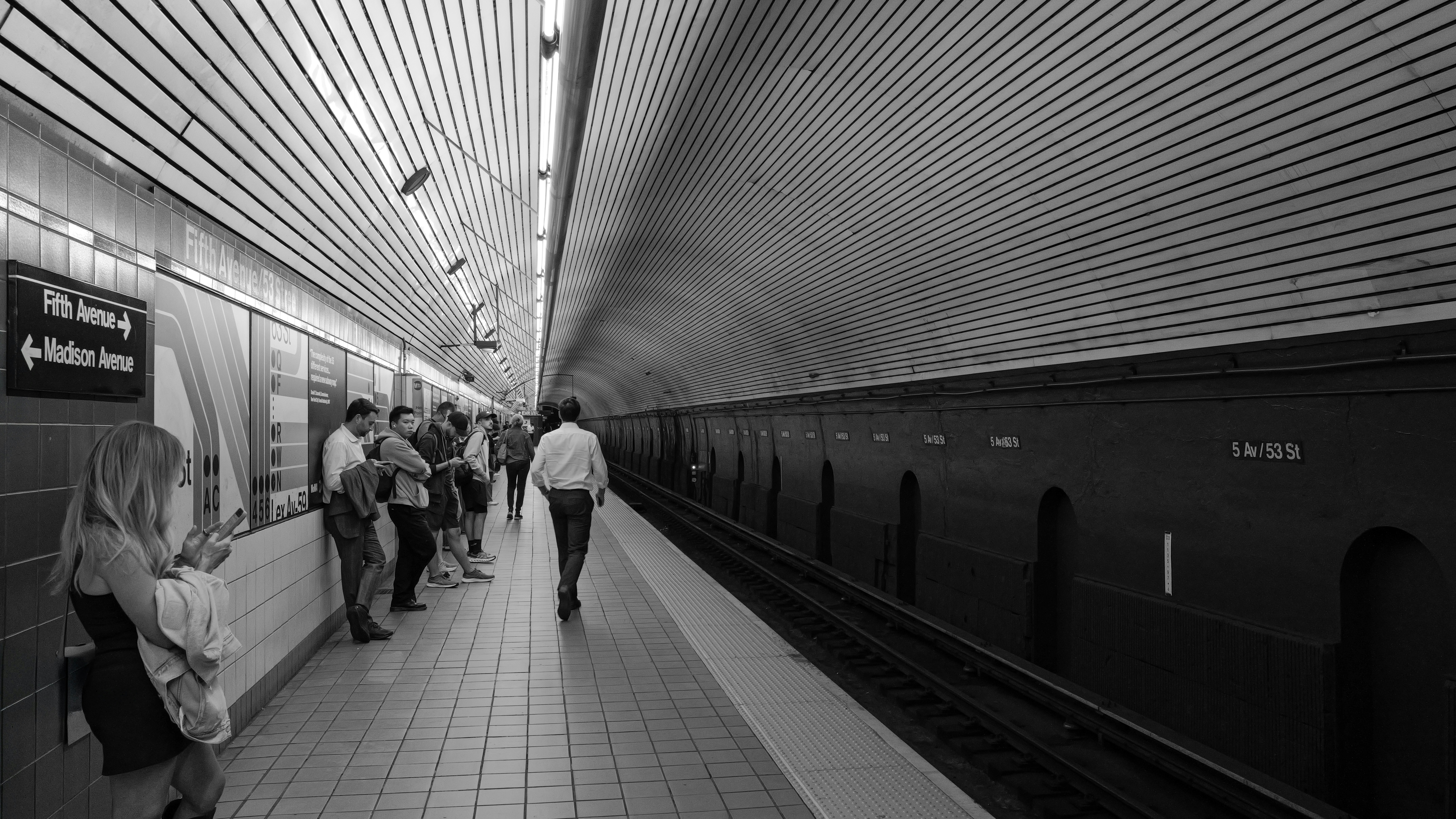 Black and white scene of a subway platform, featuring waiting commuters and a distant figure walking towards the train. The architectural lines create a sense of depth.
