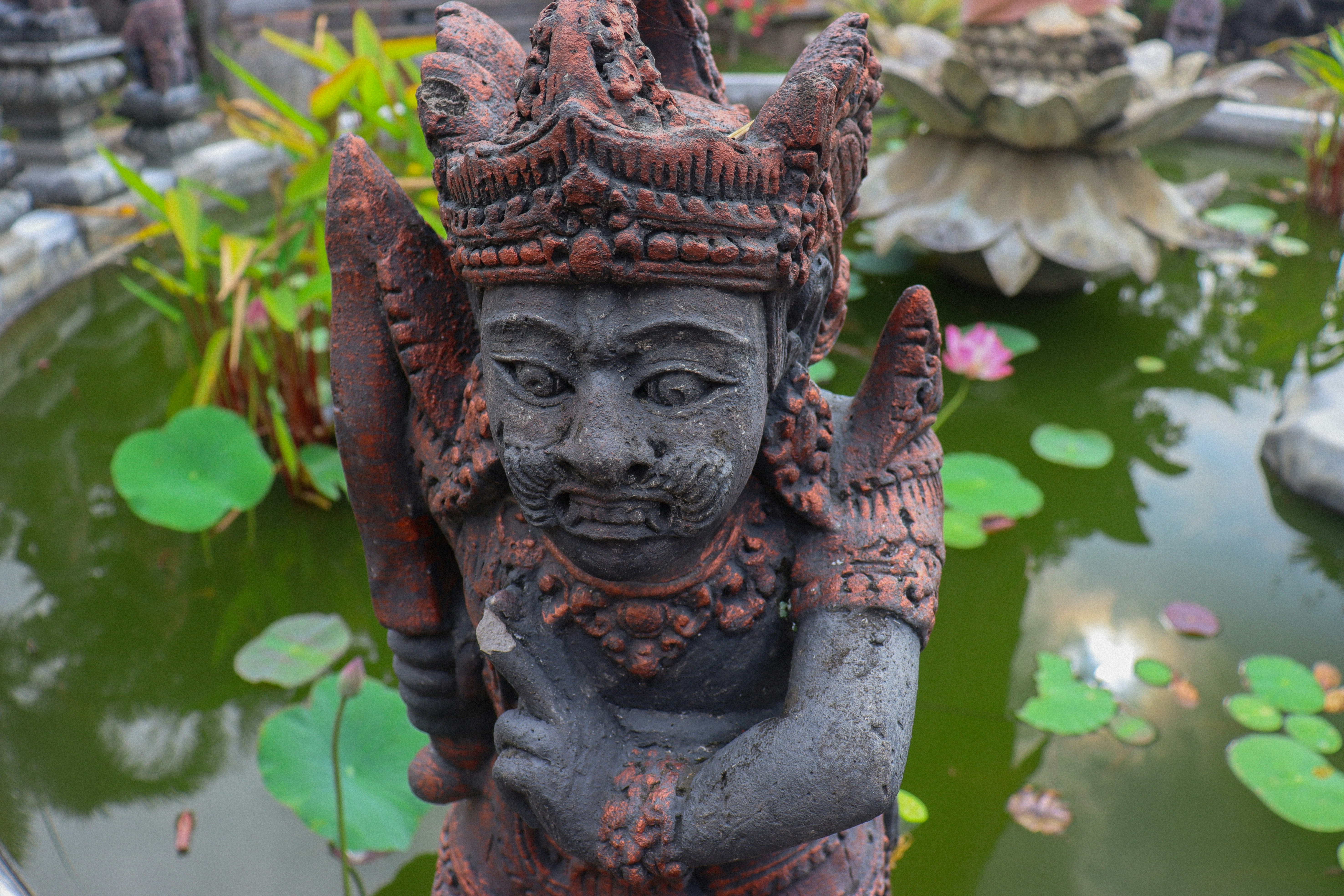 Intricately carved stone statue of a guardian figure standing amidst a tranquil pond adorned with lily pads.