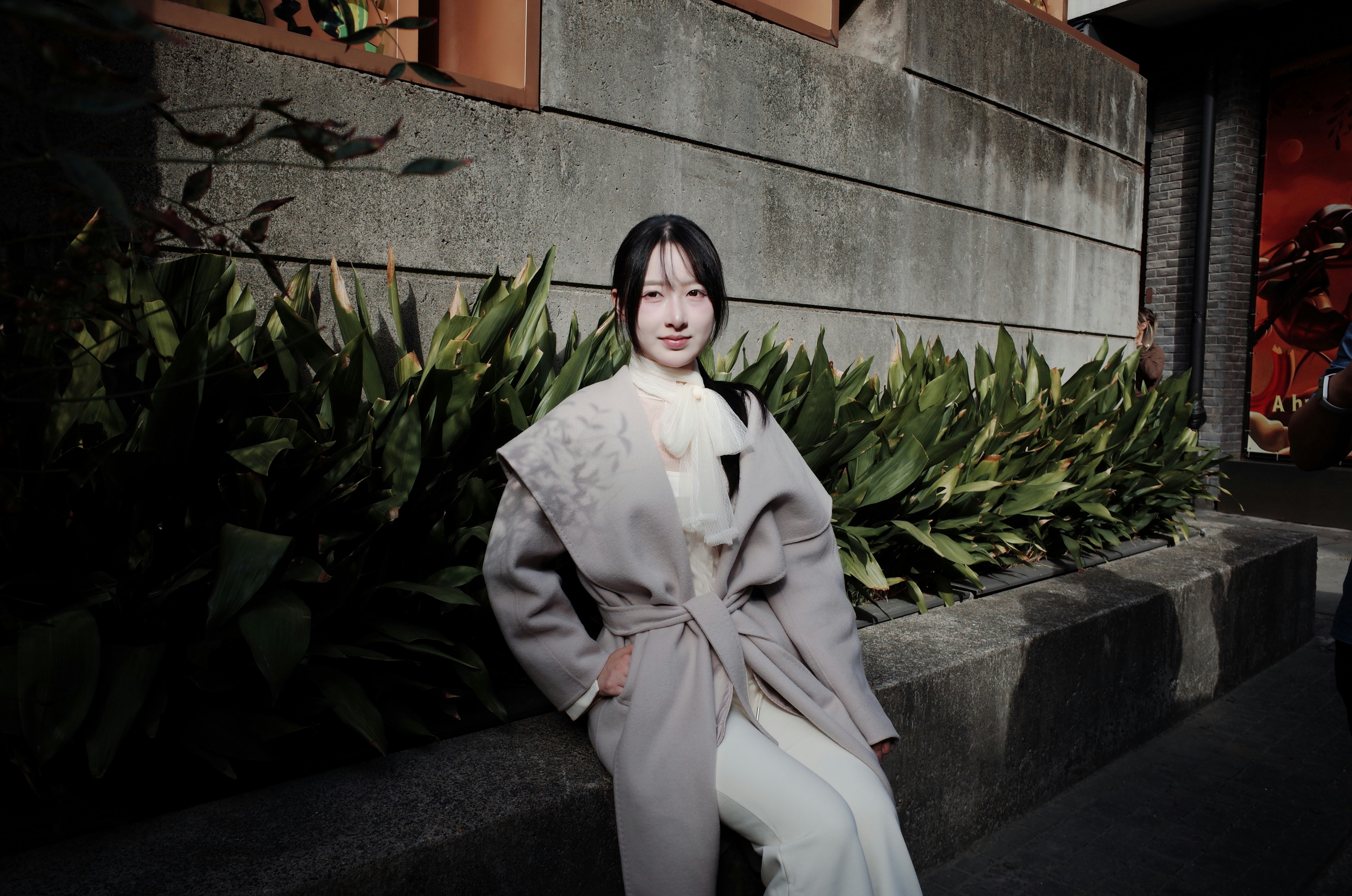 Young woman in stylish coat sitting outdoors near plants.