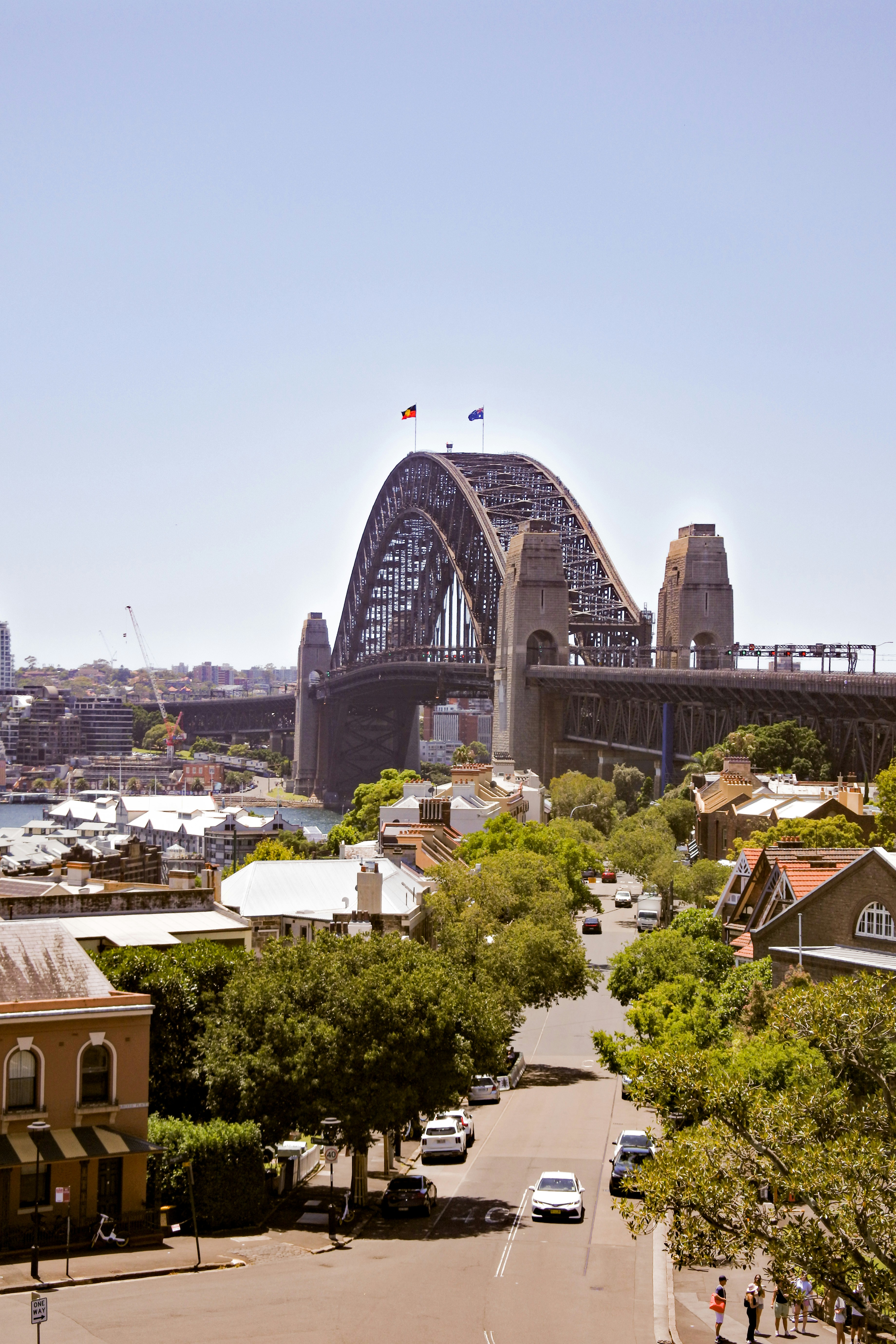 City street leading to a grand bridge, with trees lining the road and a clear sky above; a distant urban landscape beneath a prominent arch.