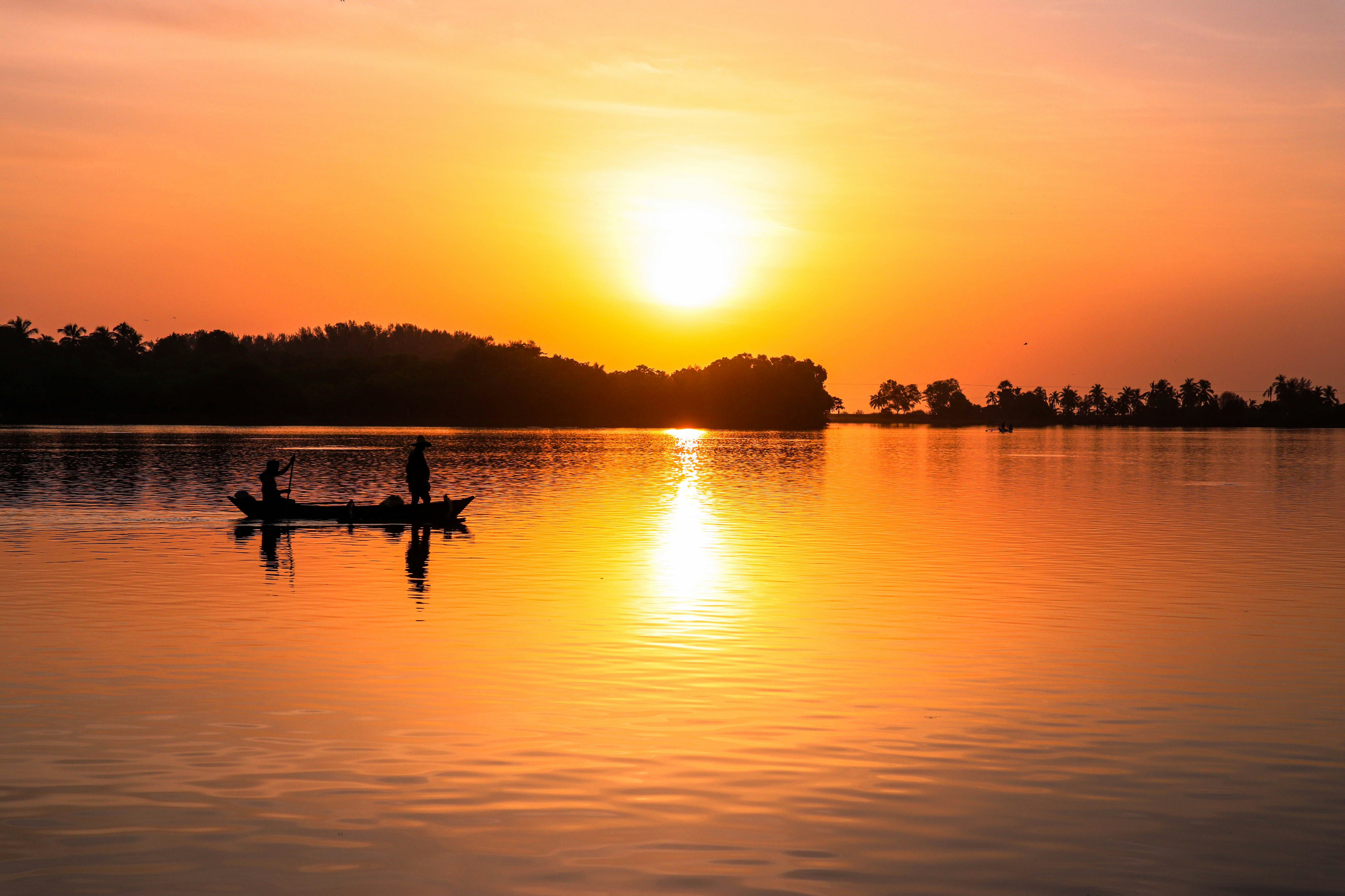 This photograph captures a breathtaking sunset over calm, reflective water, likely a lake, lagoon, or very still ocean. The entire scene is bathed in a warm, fiery orange and yellow glow from the setting sun, which creates a bright, intense sunburst at the center top. The sun's light forms a shimmering, golden reflection on the water's surface, leading from the horizon toward the foreground. In the middle ground, a silhouette of a small fishing boat or canoe with two people aboard is visible against the bright light. The horizon is marked by a dark silhouette of lush trees and tropical foliage | Two people in a boat on a lake at sunset.