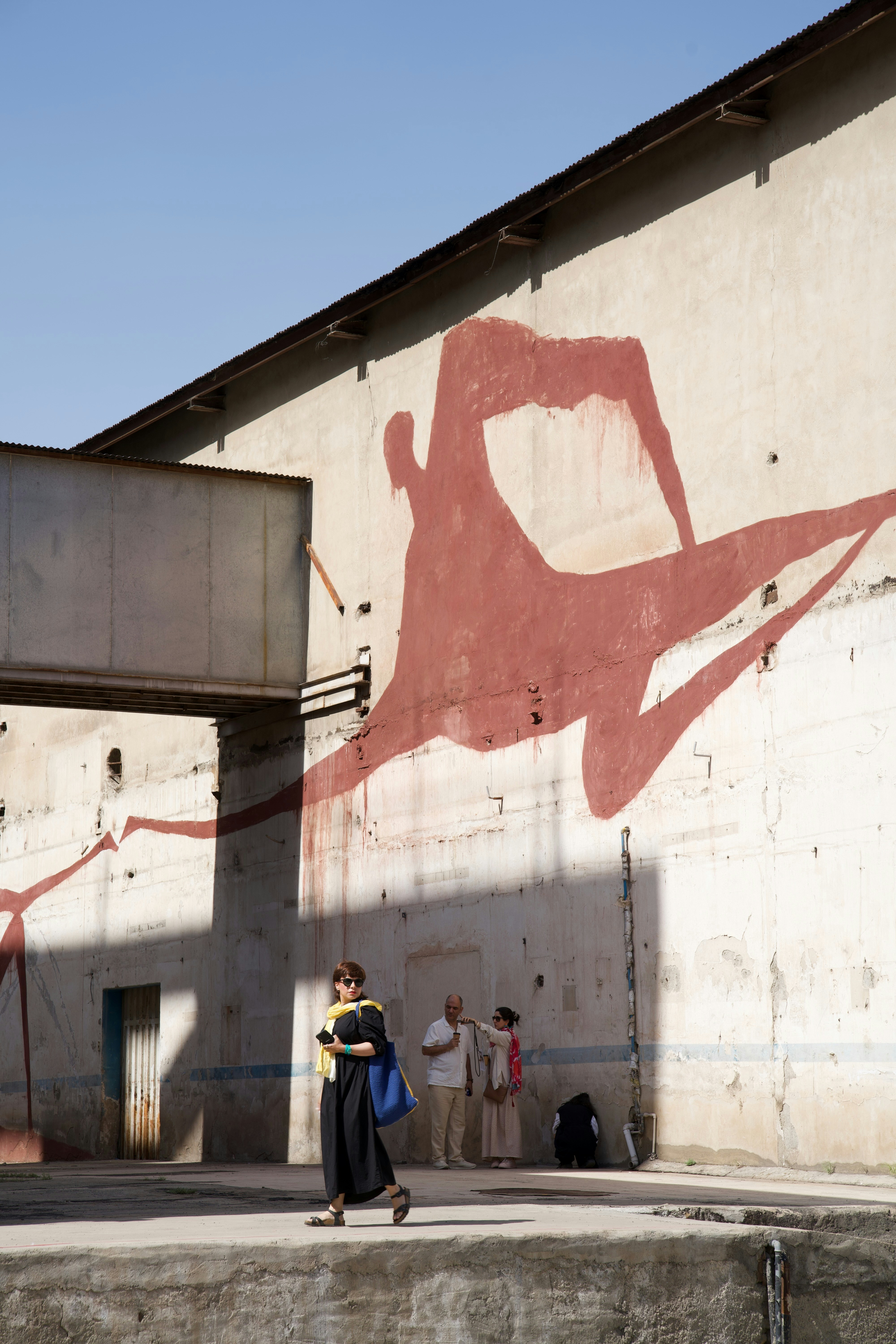 Mirza Hamid has been painting familiar motifs on the walls of Tehran for years, using only a single color. He prefers to remain anonymous, earning the title of the "Poet of Tehran's Walls." His works consistently feature figures, relationships, and symbols that explore the timeless concepts of human existence. For the first time, he has displayed his artwork on the walls of abandoned warehouses left over from an old refrigerator factory in Tehran, showcasing his creations without being physically present. | Woman walks past mural on building wall